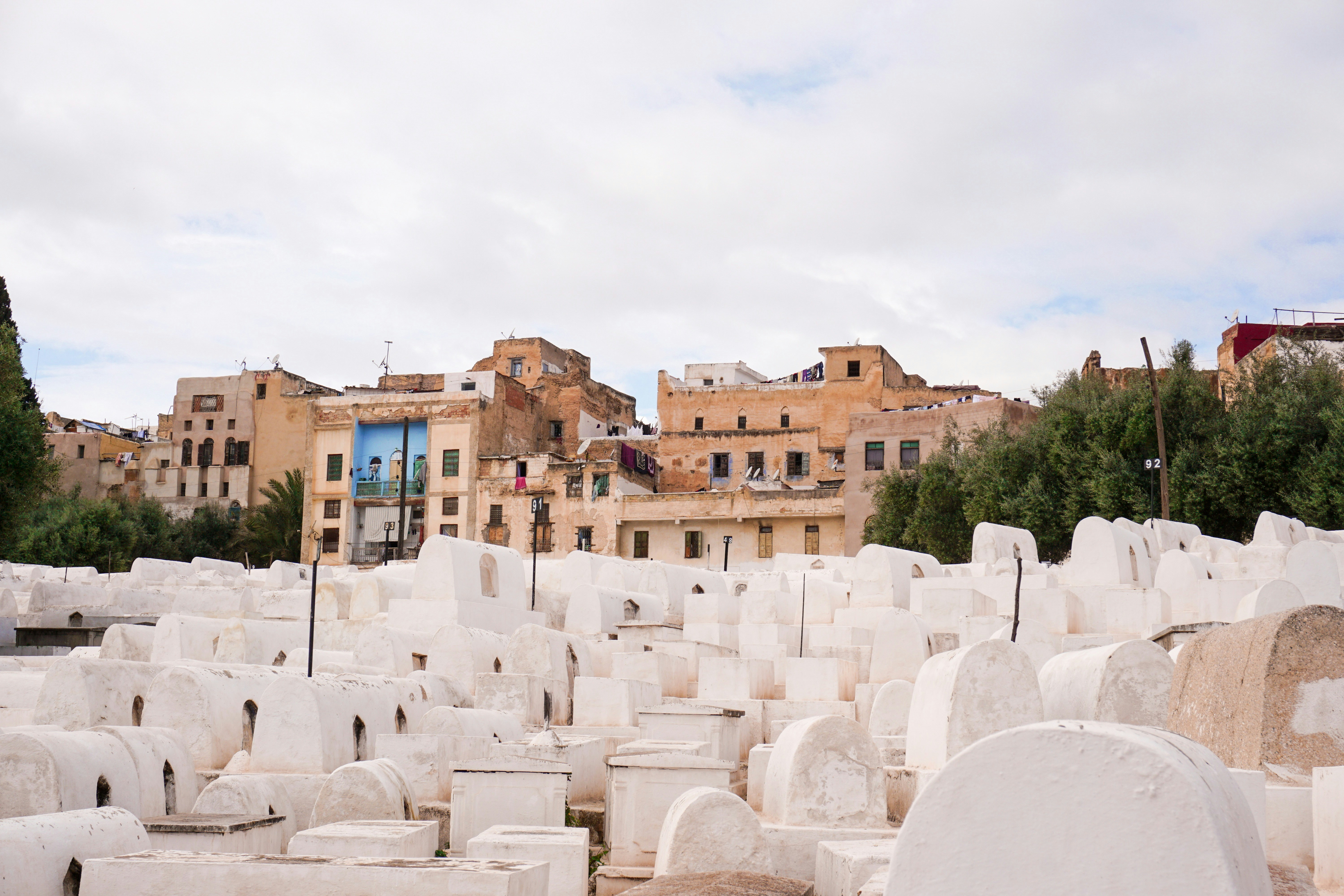 White tombstones of a Jewish cemetery with Moroccan houses in the background under a cloudy sky.