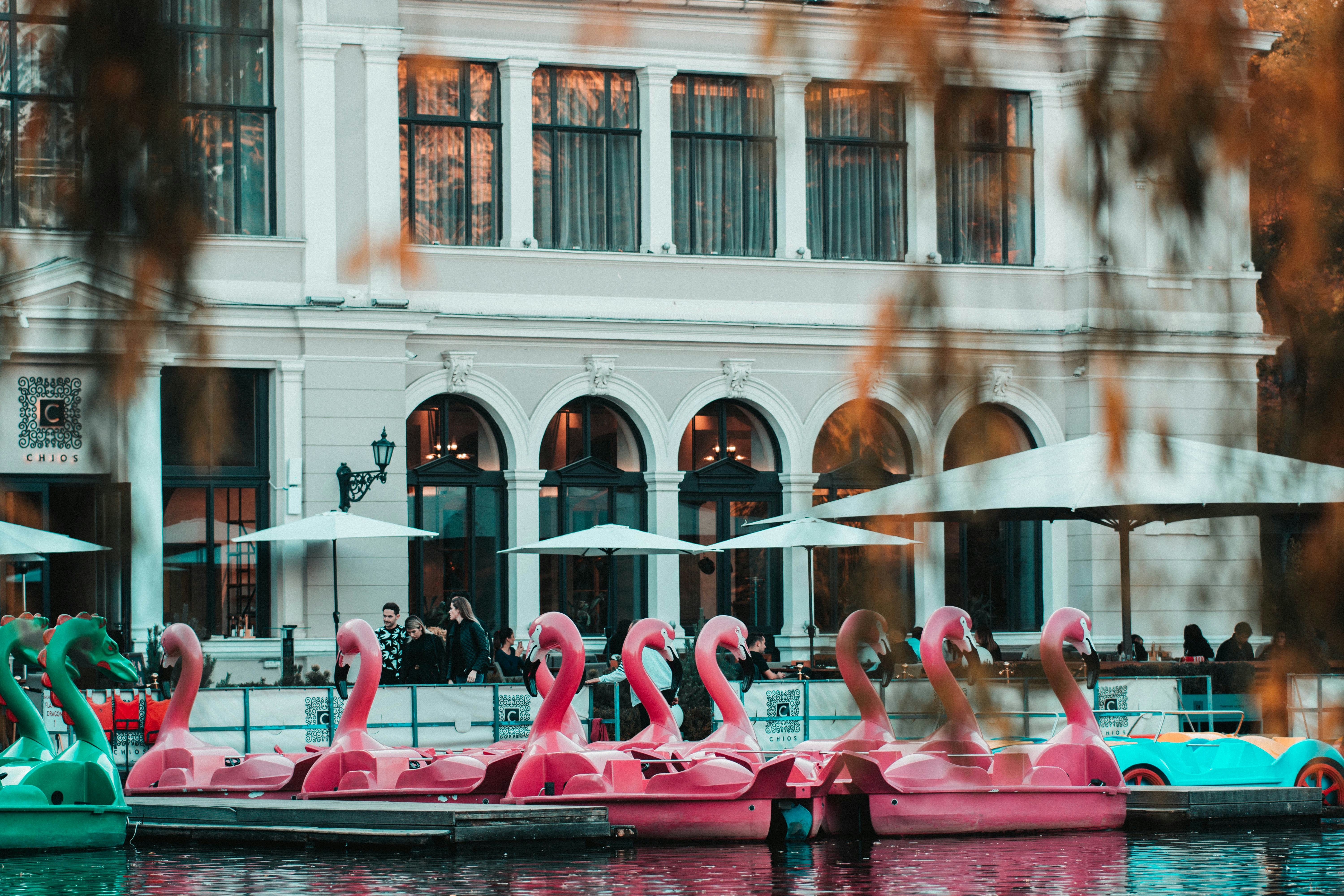Pink and green flamingo paddle boats float beside a riverside café with large arched windows.