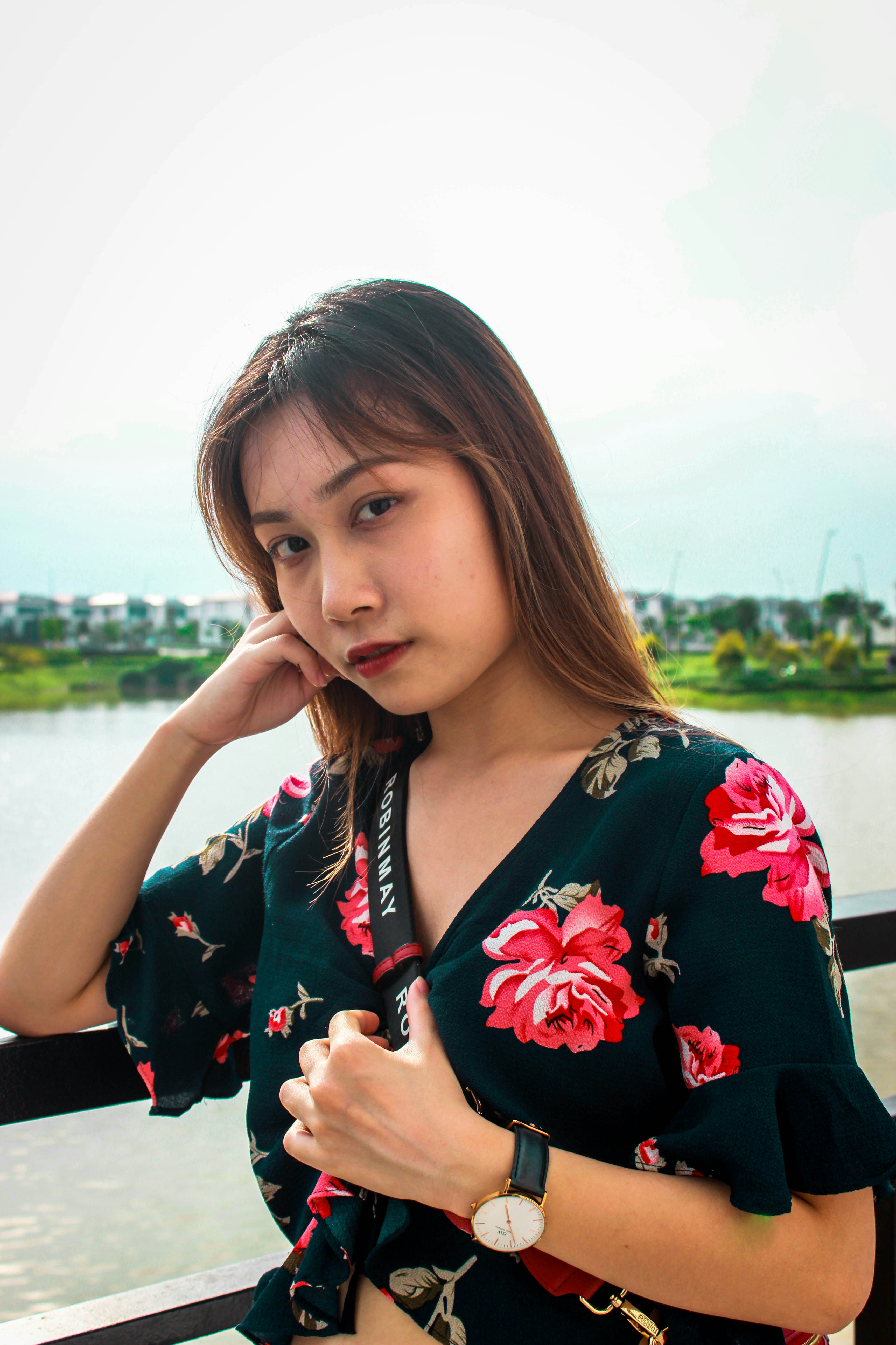 Young woman posing thoughtfully against a backdrop of water and greenery, wearing a floral blouse and a watch.