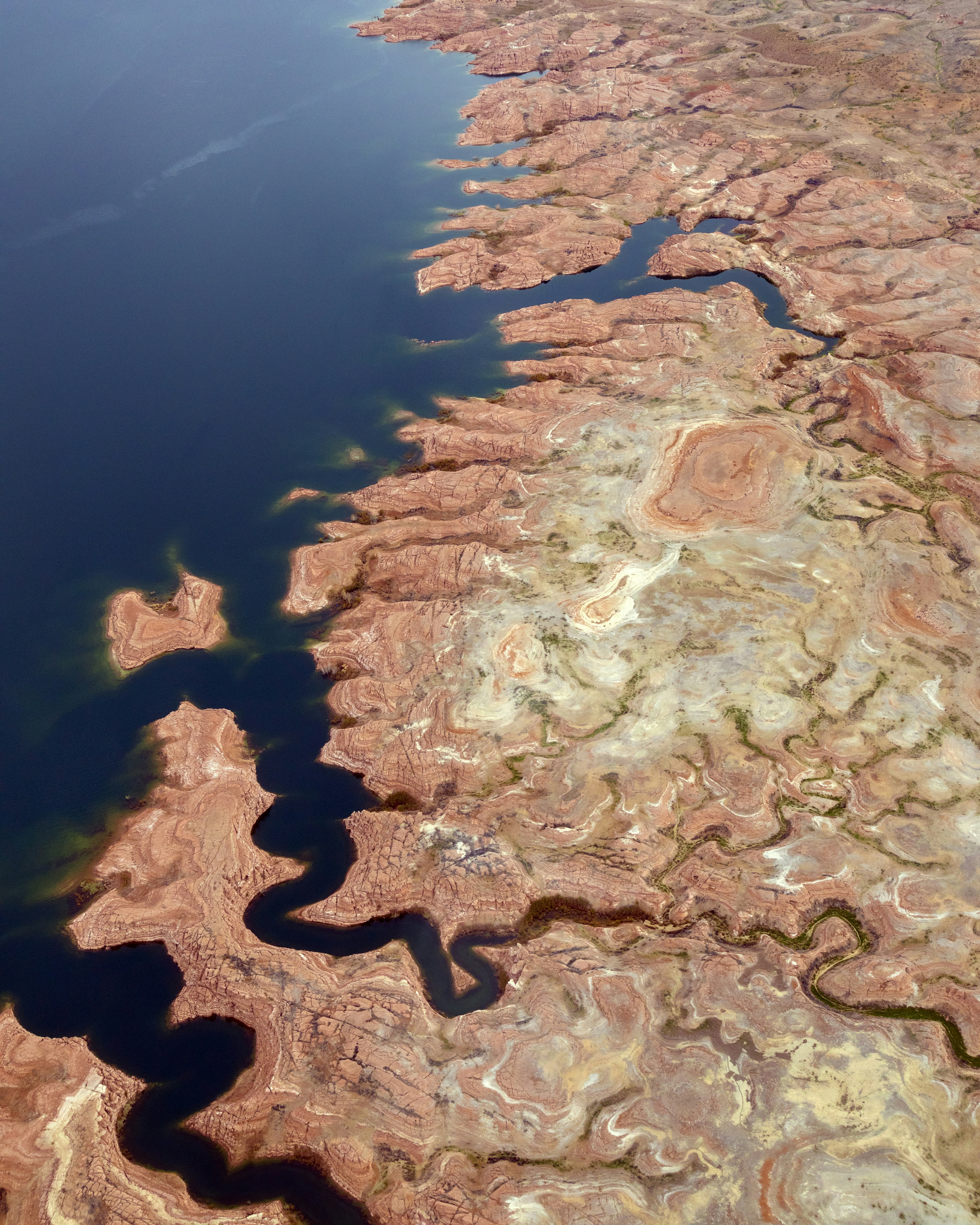 Aerial view of a winding shoreline where vibrant earth tones meet the deep blue of a lake, showcasing the intricate patterns of the landscape.