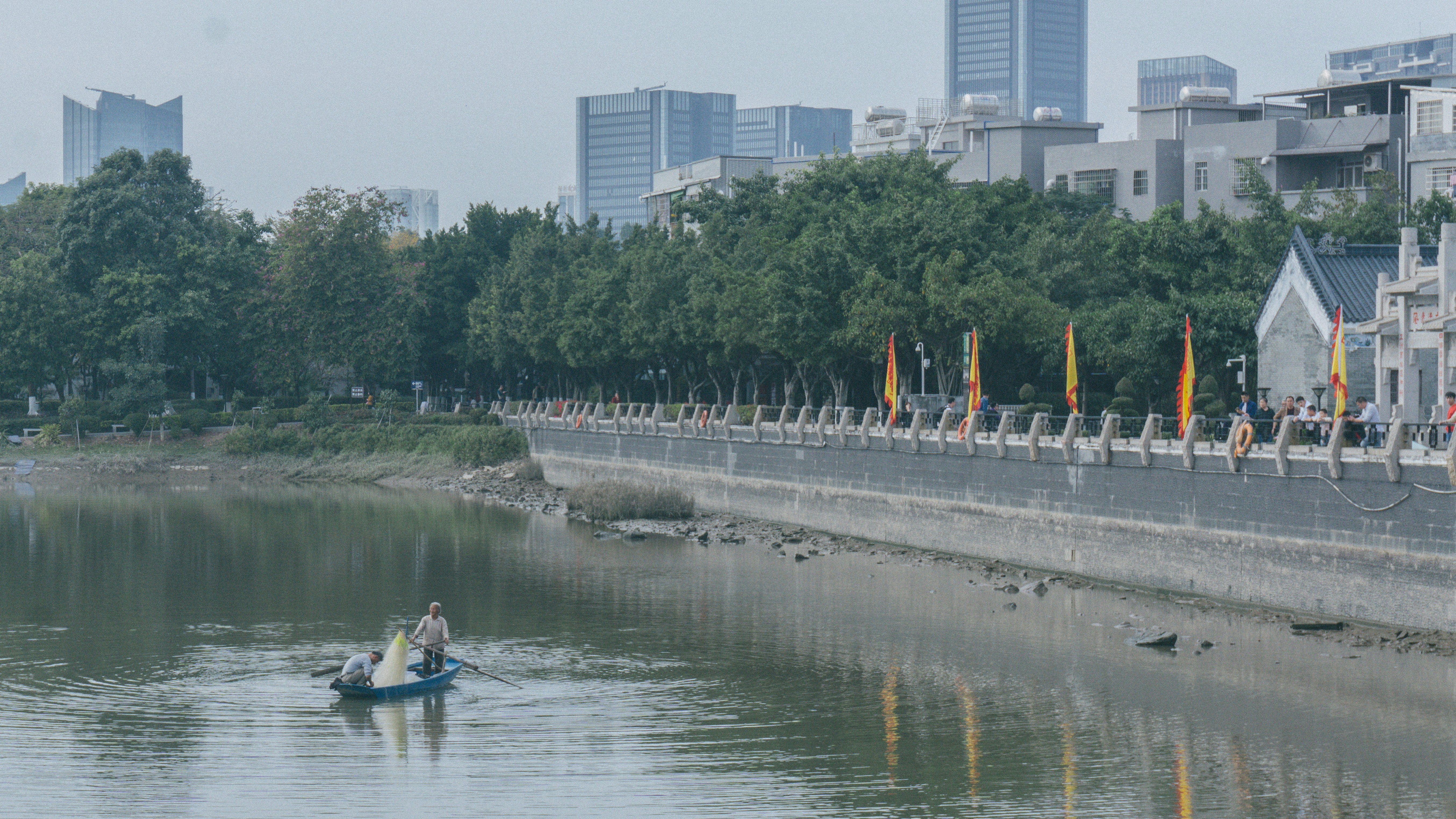 A photograph of a small motorboat on a calm river beside a tree-lined promenade, with flags along the railing and modern buildings in the background.