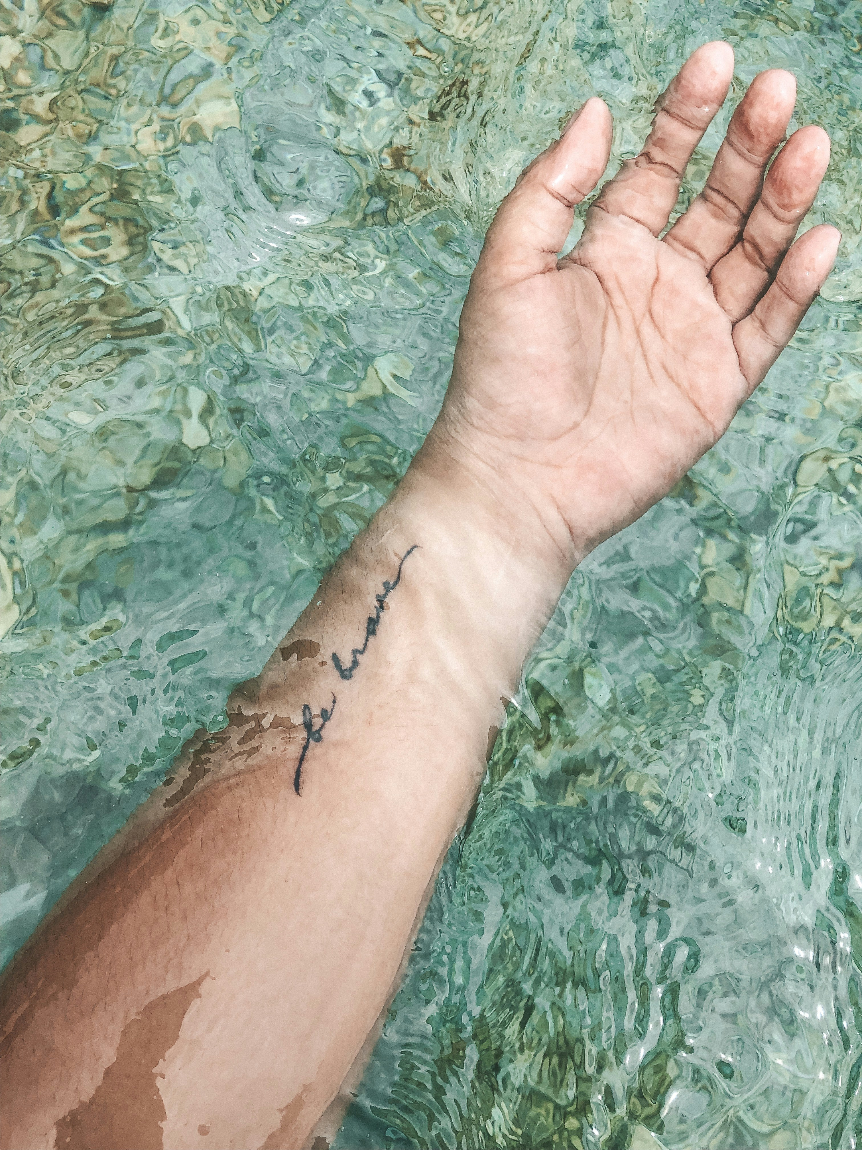A hand submerged in clear water, showcasing a delicate tattoo on the wrist. The fluidity of the water enhances the tattoo's intricate design.