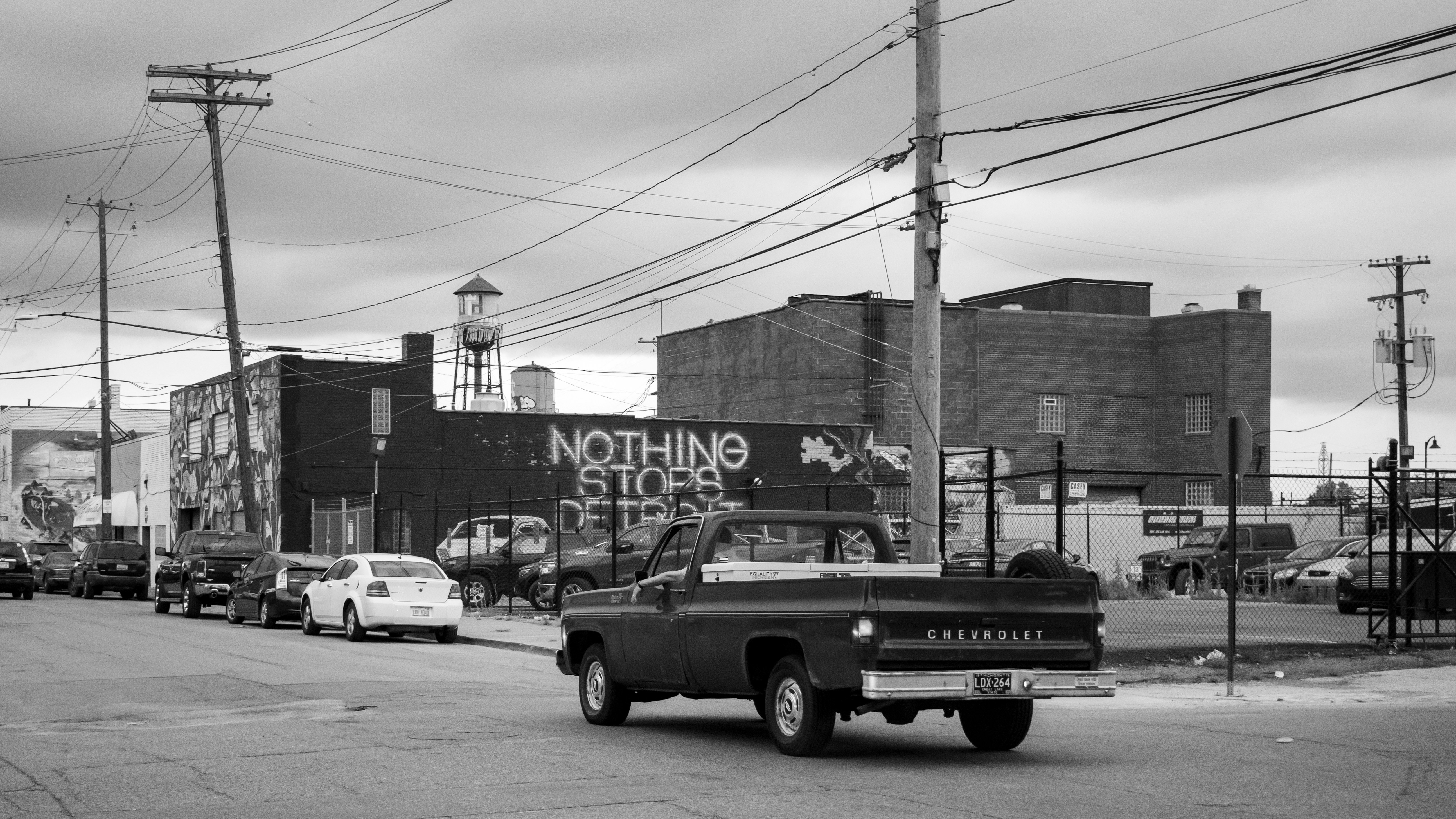 Grayscale scene of a man driving a single cab pickup truck down a city street lined with industrial buildings.