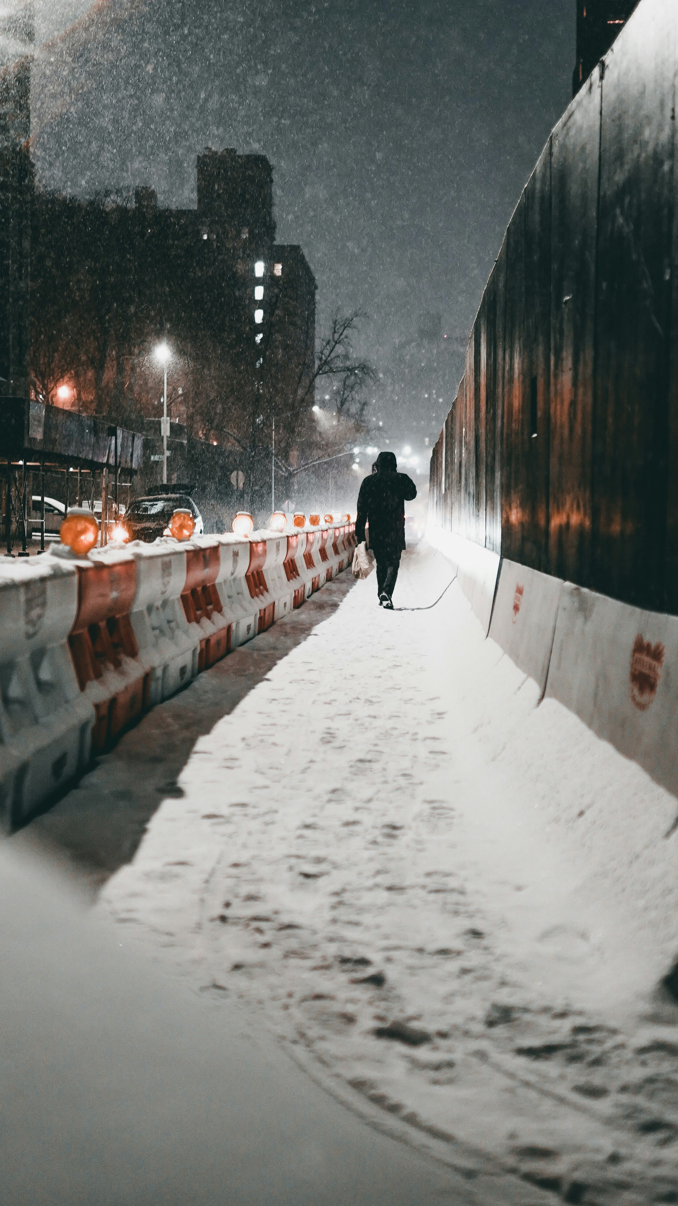 A solitary figure walks along a snow-covered path lined with construction barriers, illuminated by distant city lights during a snowstorm.