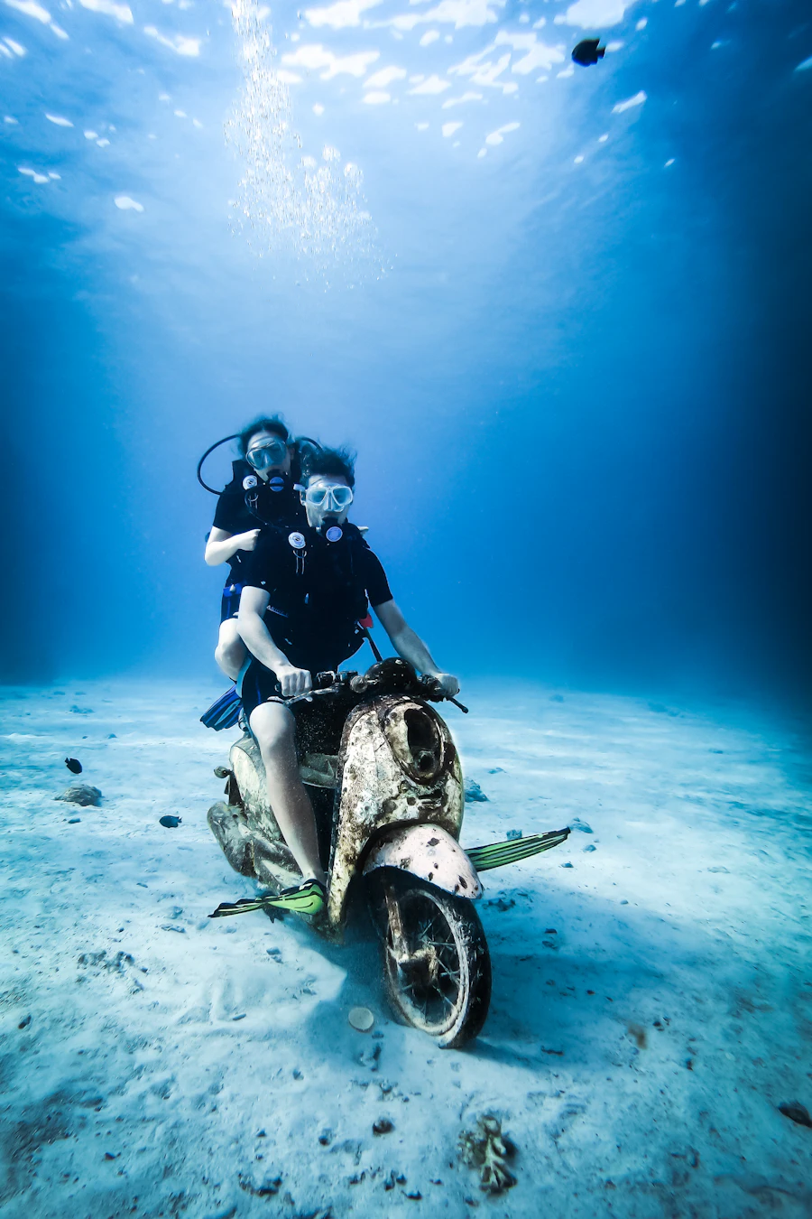 Two scuba divers sit on a coral-encrusted scooter on the seafloor, with clear blue water and sunlight filtering from above.