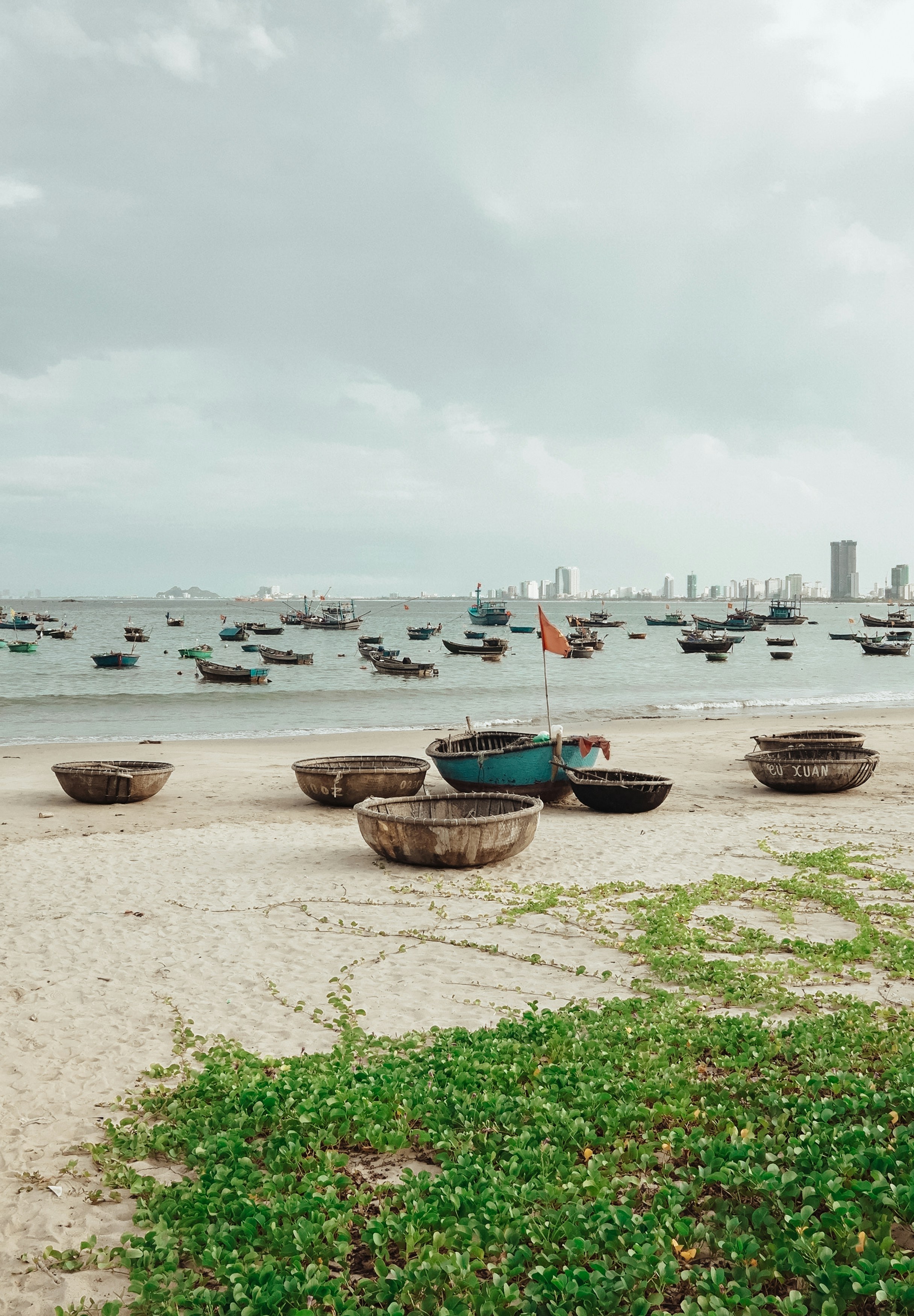 Traditional round fishing boats anchored near the shore, with a backdrop of a bustling city skyline under a cloudy sky.