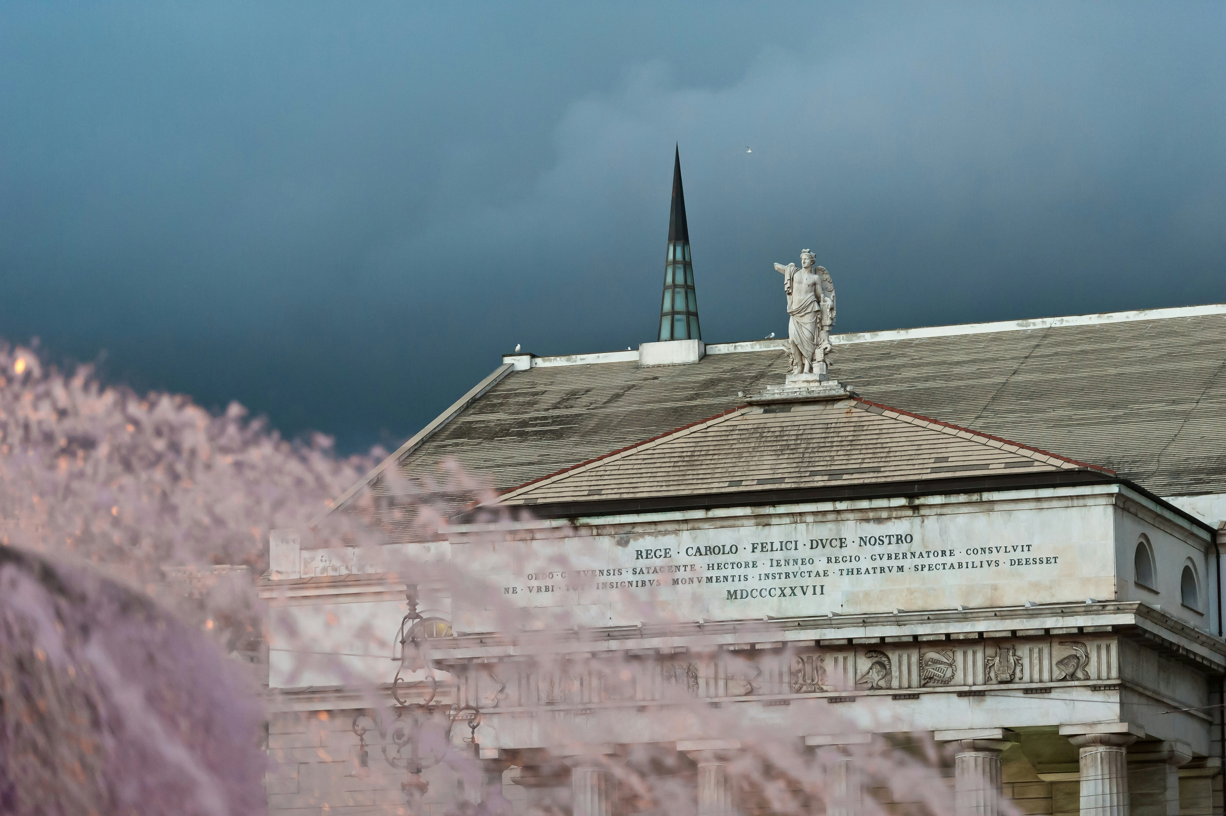 Historic building topped with a statue, framed by a dramatic dark sky and foreground fountain.