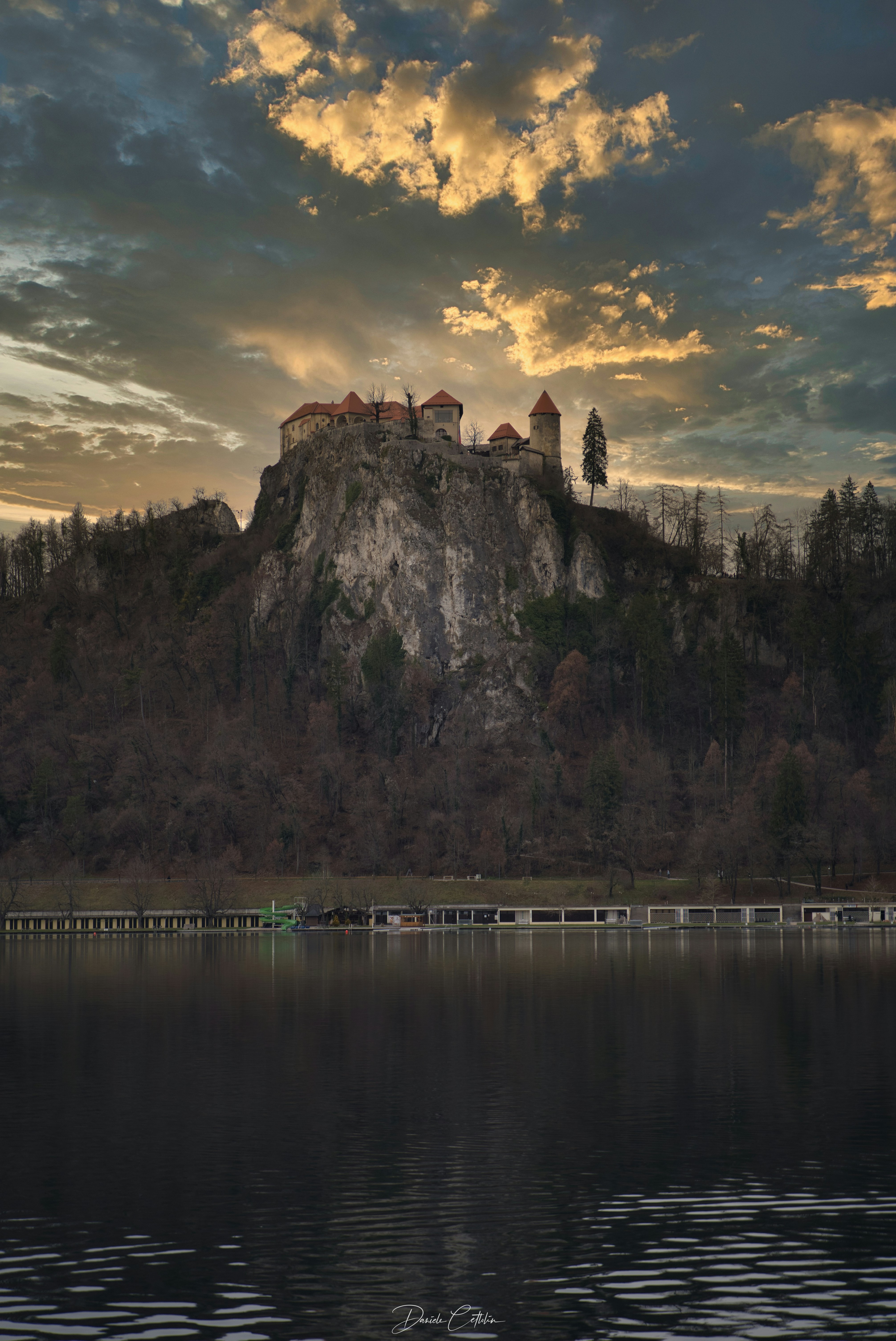 A medieval castle perched atop a rocky cliff, surrounded by tranquil waters reflecting the dramatic sky at dusk.