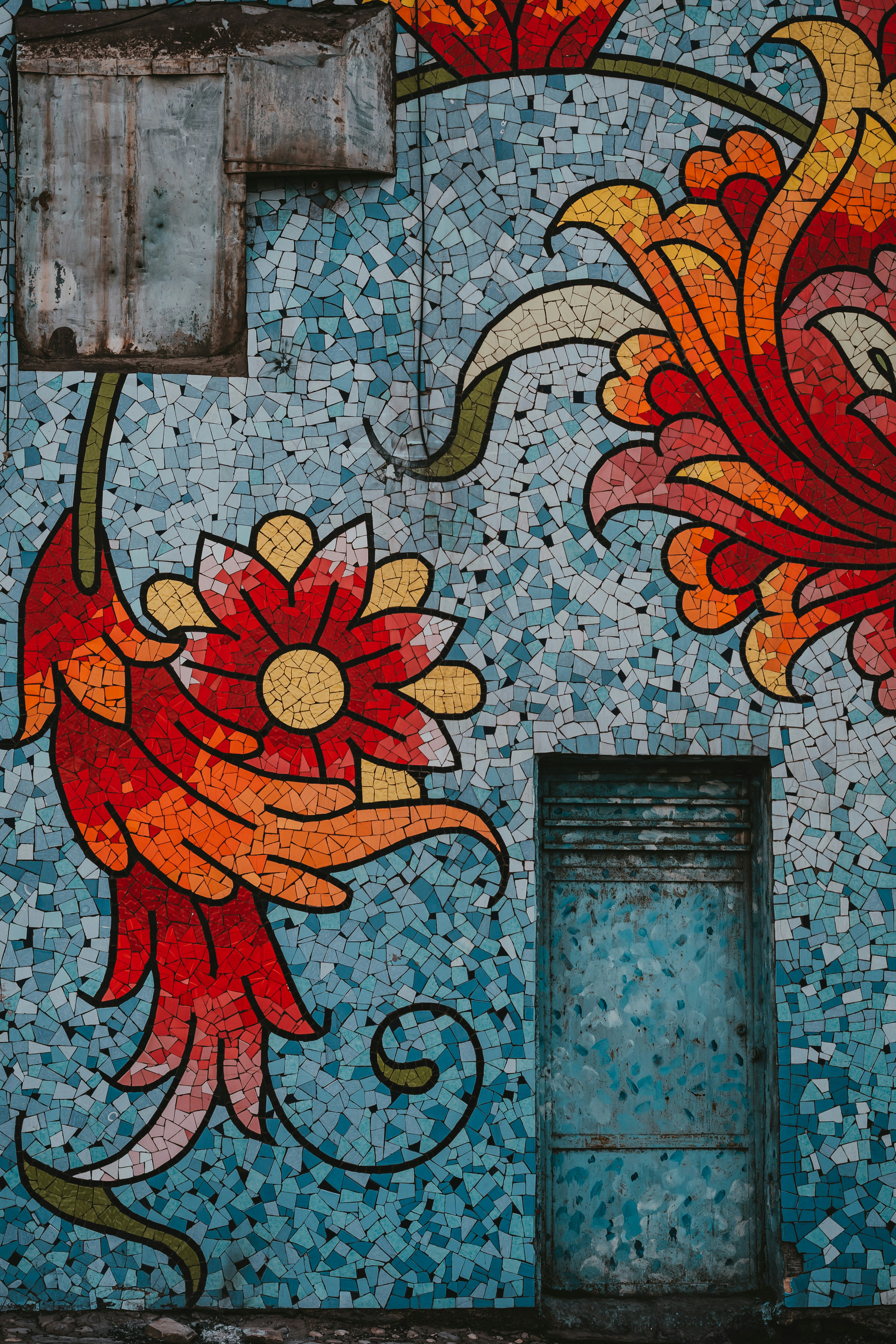 Colorful mosaic mural featuring floral designs against a blue background, with a weathered door integrated into the artwork.