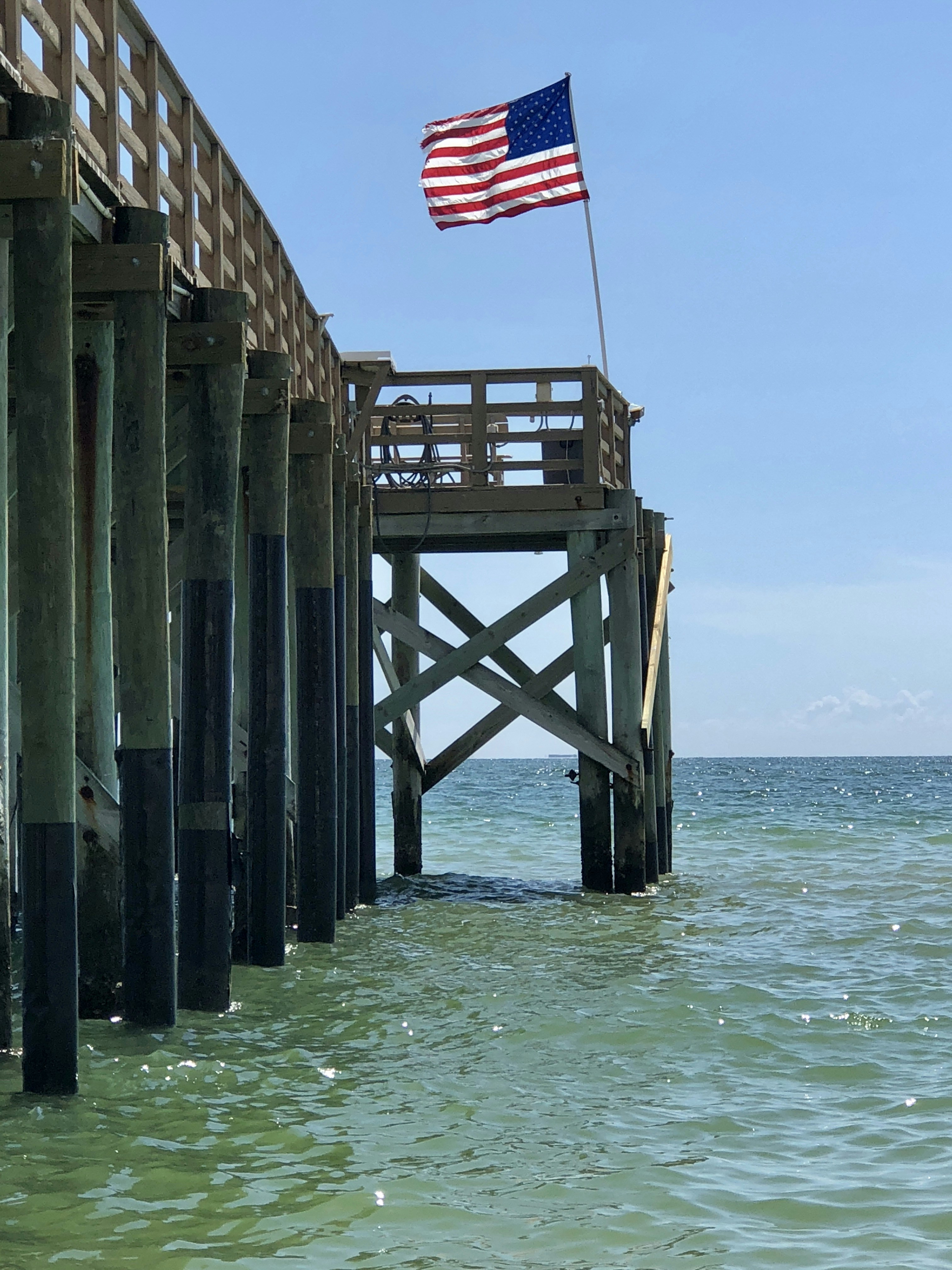 American flag waving atop a wooden pier, surrounded by gentle ocean waves under a clear blue sky.