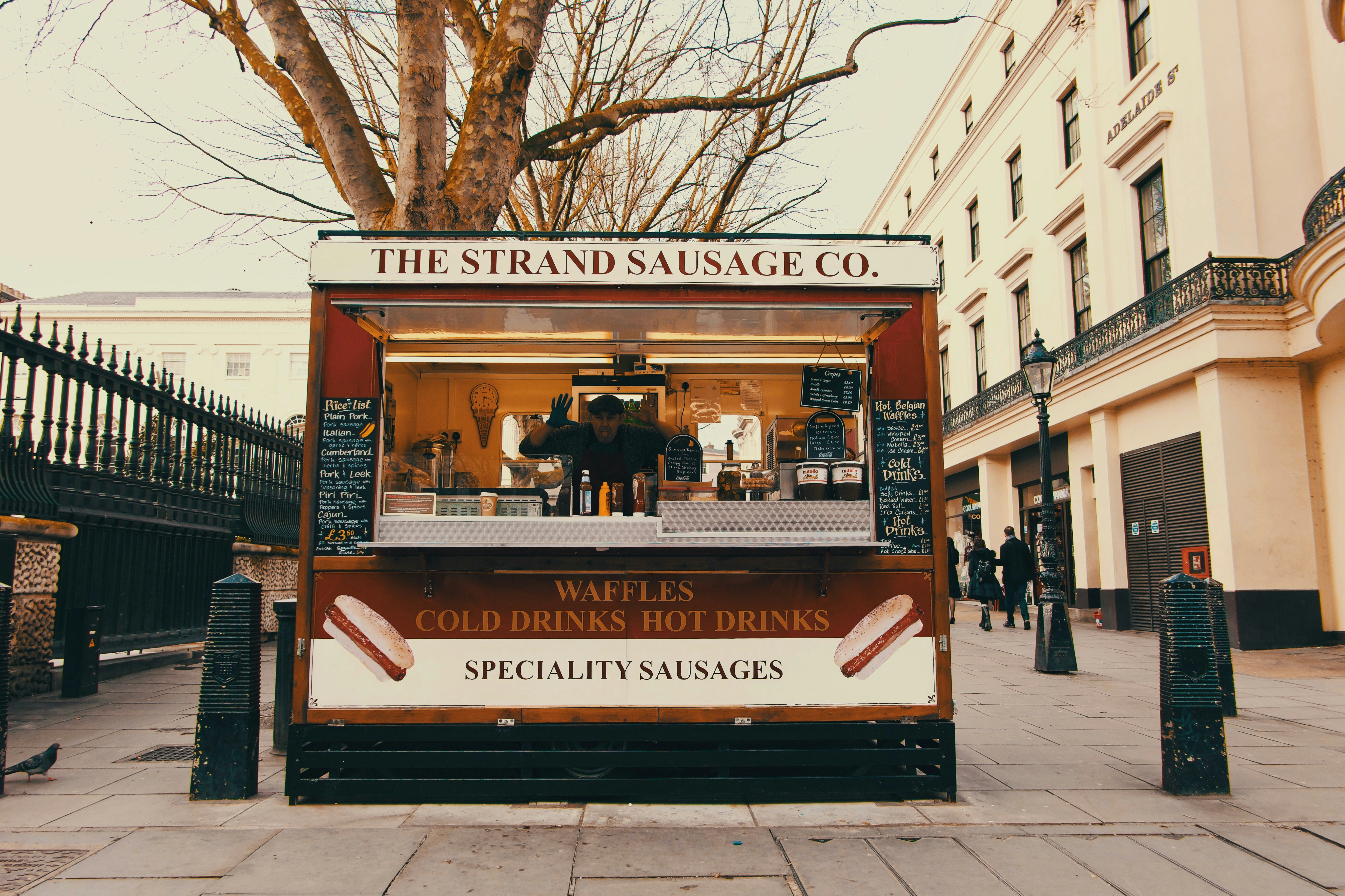 Photograph of a sausage cart labeled The Strand Sausage Co. on a quiet city street, with signage advertising waffles and drinks.