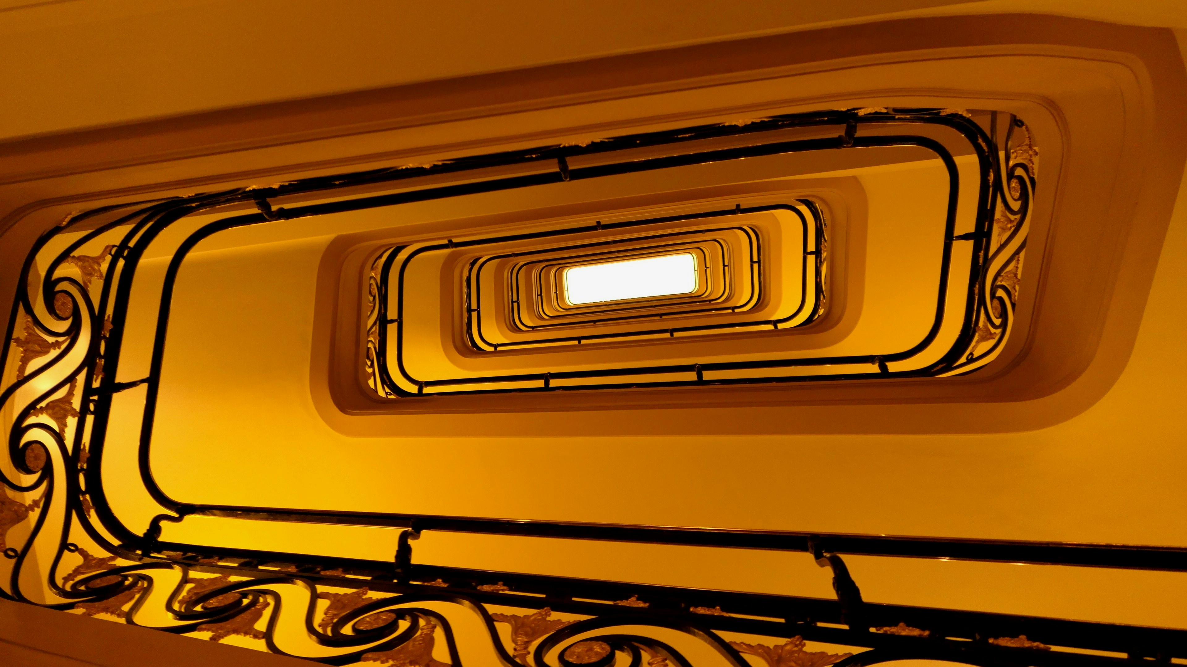 An architectural photograph of a spiraling stairwell bathed in warm amber light, emphasizing the repeating black rails and layered landings. The bright rectangular opening at the top serves as the visual anchor.