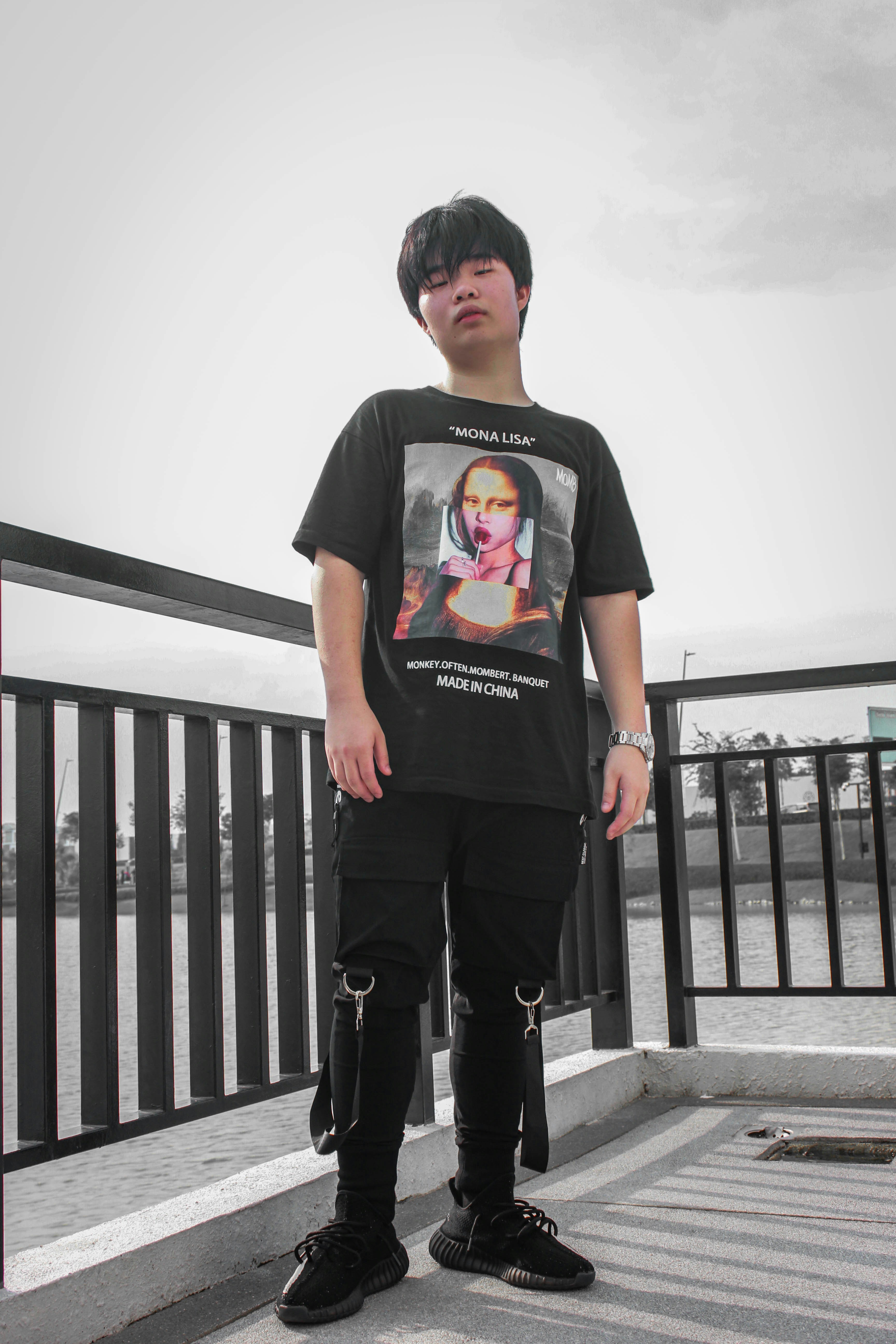 Young person standing confidently against a railing, wearing a graphic tee featuring a modern interpretation of the Mona Lisa. Urban backdrop with water in the distance.