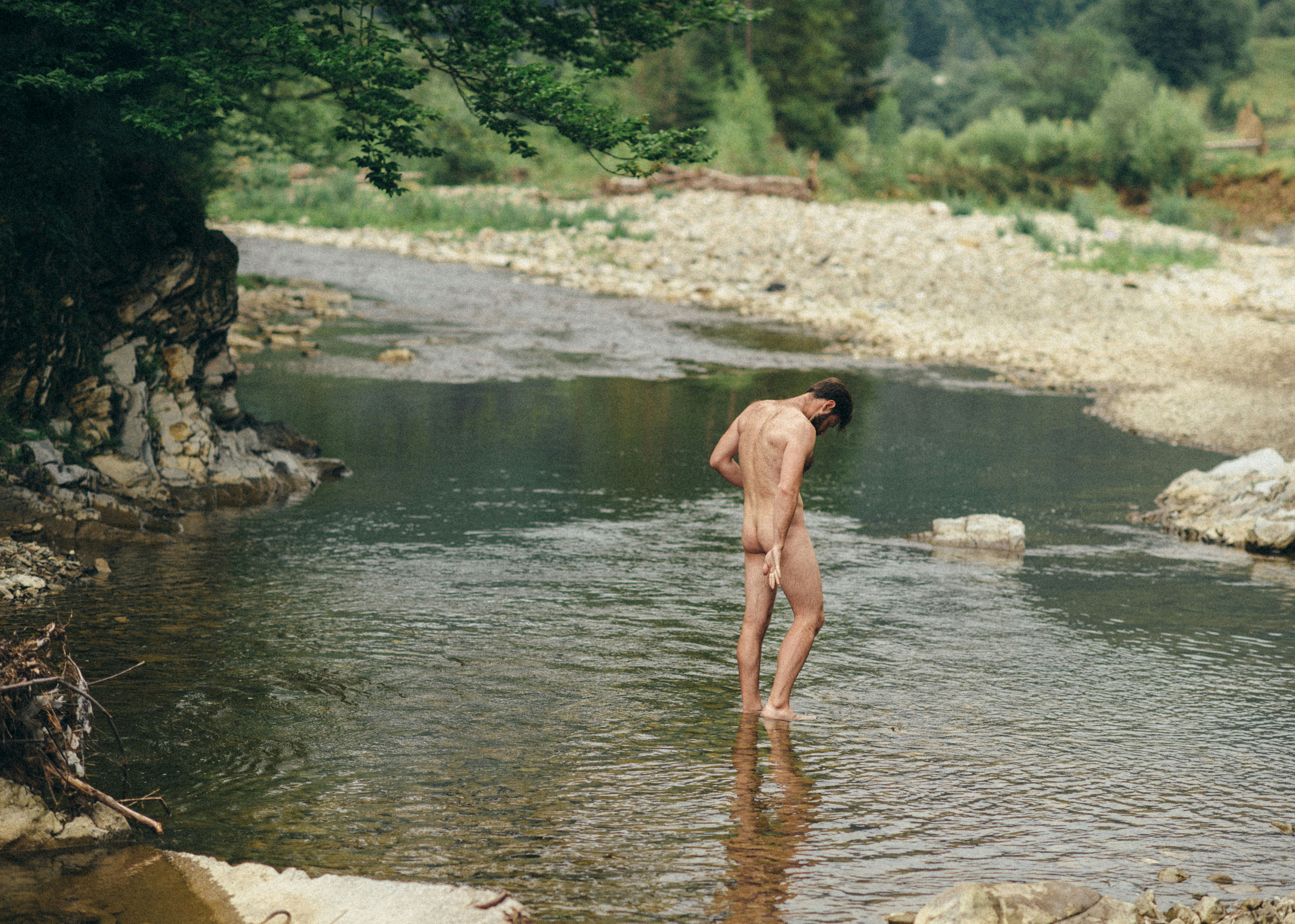 Nude figure stands barefoot in a shallow river among rocks with a forest backdrop. This photograph uses natural light and reflective water to emphasize form and texture.