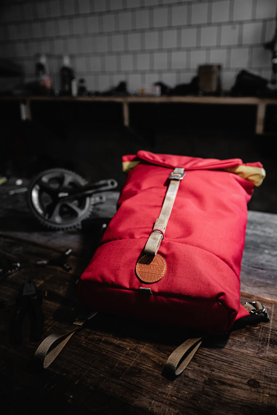 Red roll-top backpack with tan straps resting on a dark wooden table in a dim workshop. A bicycle crank and tools sit out of focus in the background.