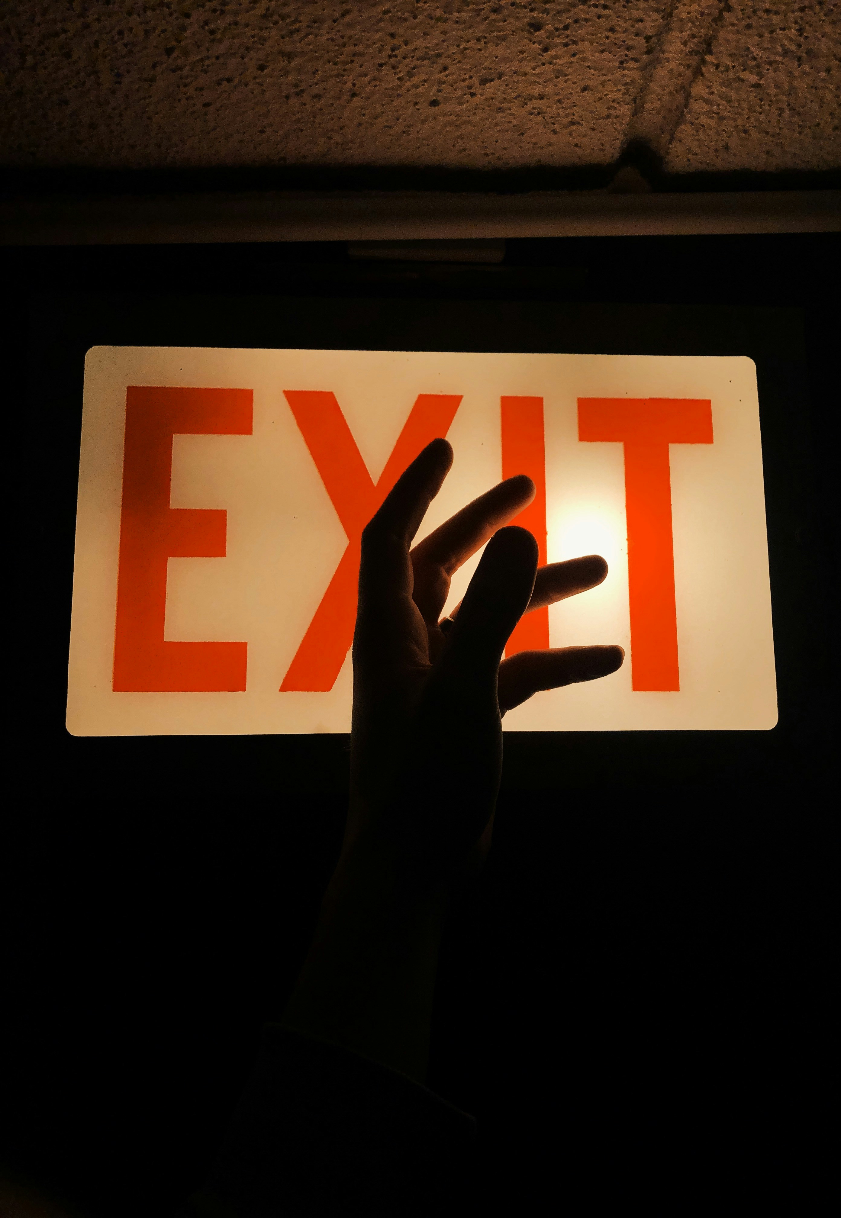 A hand reaches towards a brightly illuminated 'EXIT' sign against a dark background.
