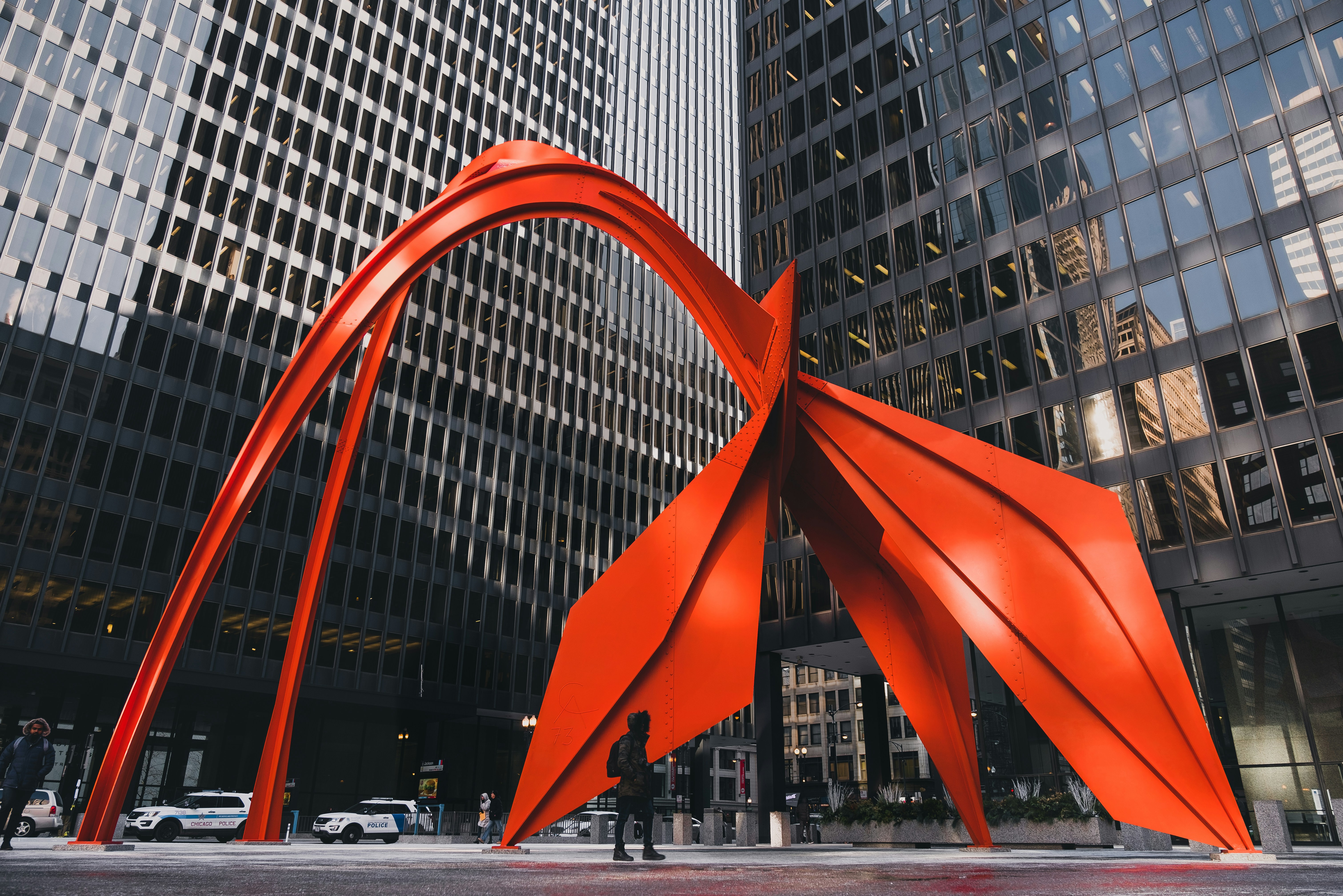 Large red metal sculpture arches in a bustling city plaza surrounded by skyscrapers.
