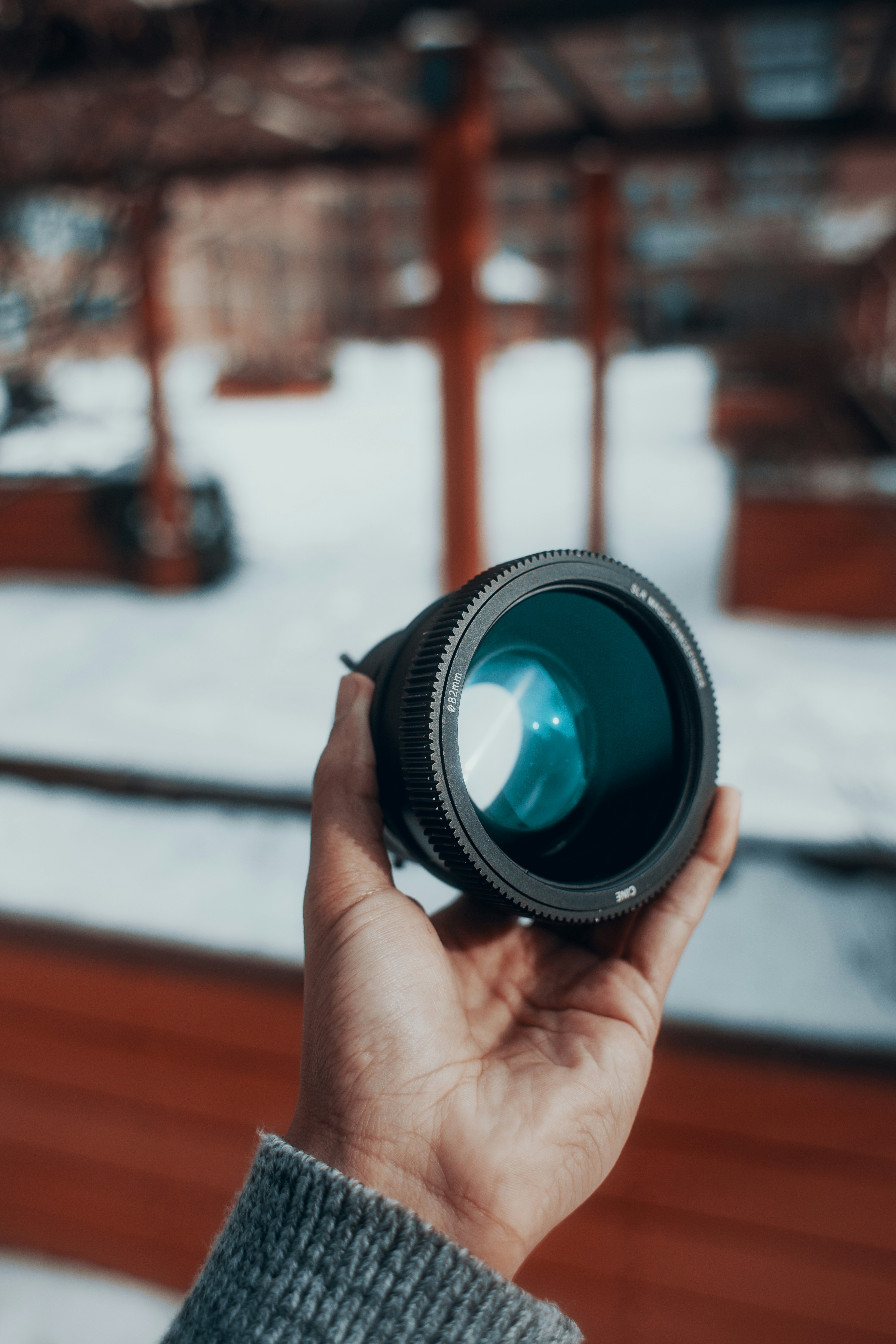 Person holding a black camera lens against a snowy outdoor backdrop with blurred wooden structures.