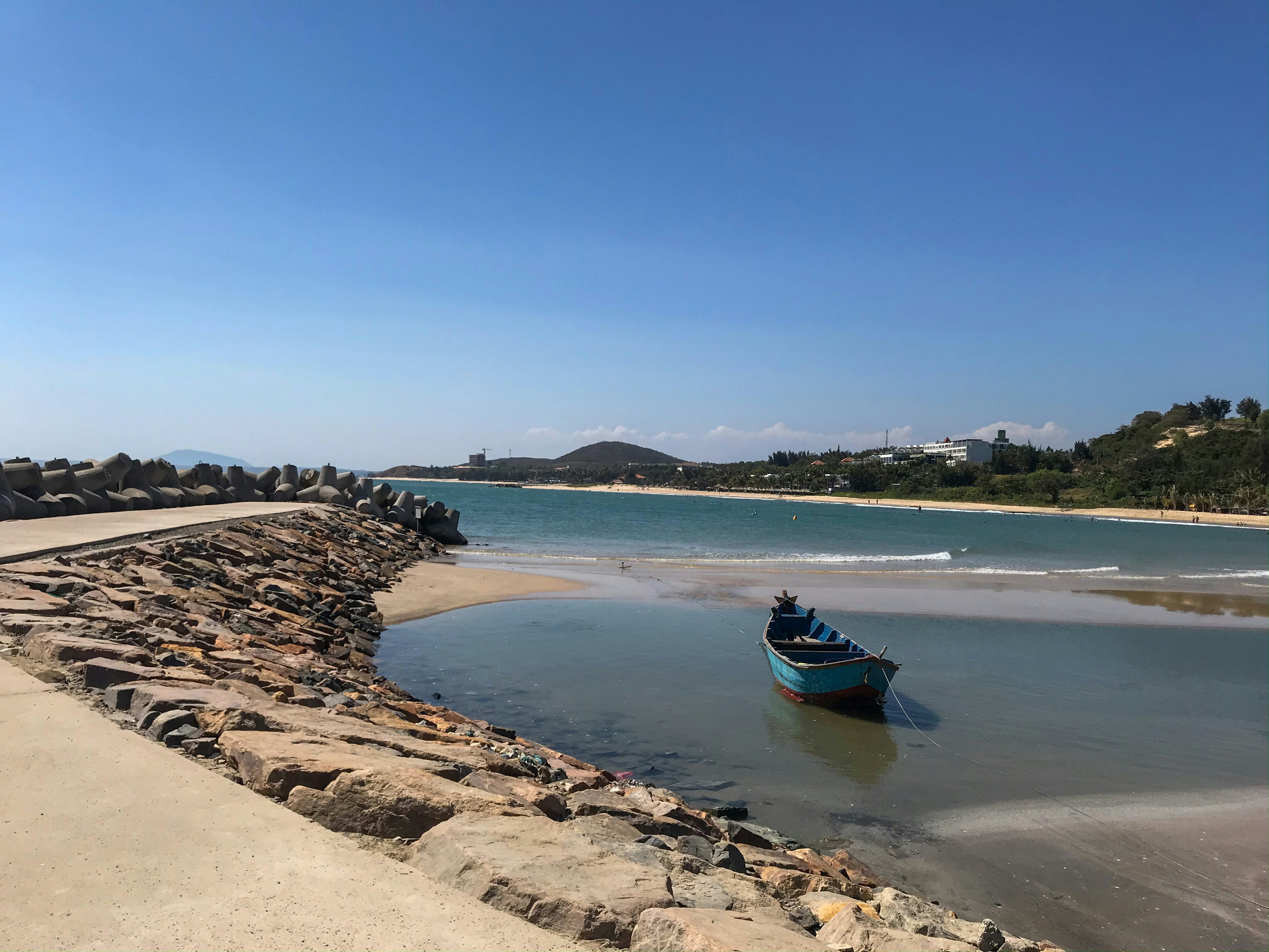 A solitary blue boat rests in shallow waters, framed by rocky shores and distant hills under a clear blue sky.