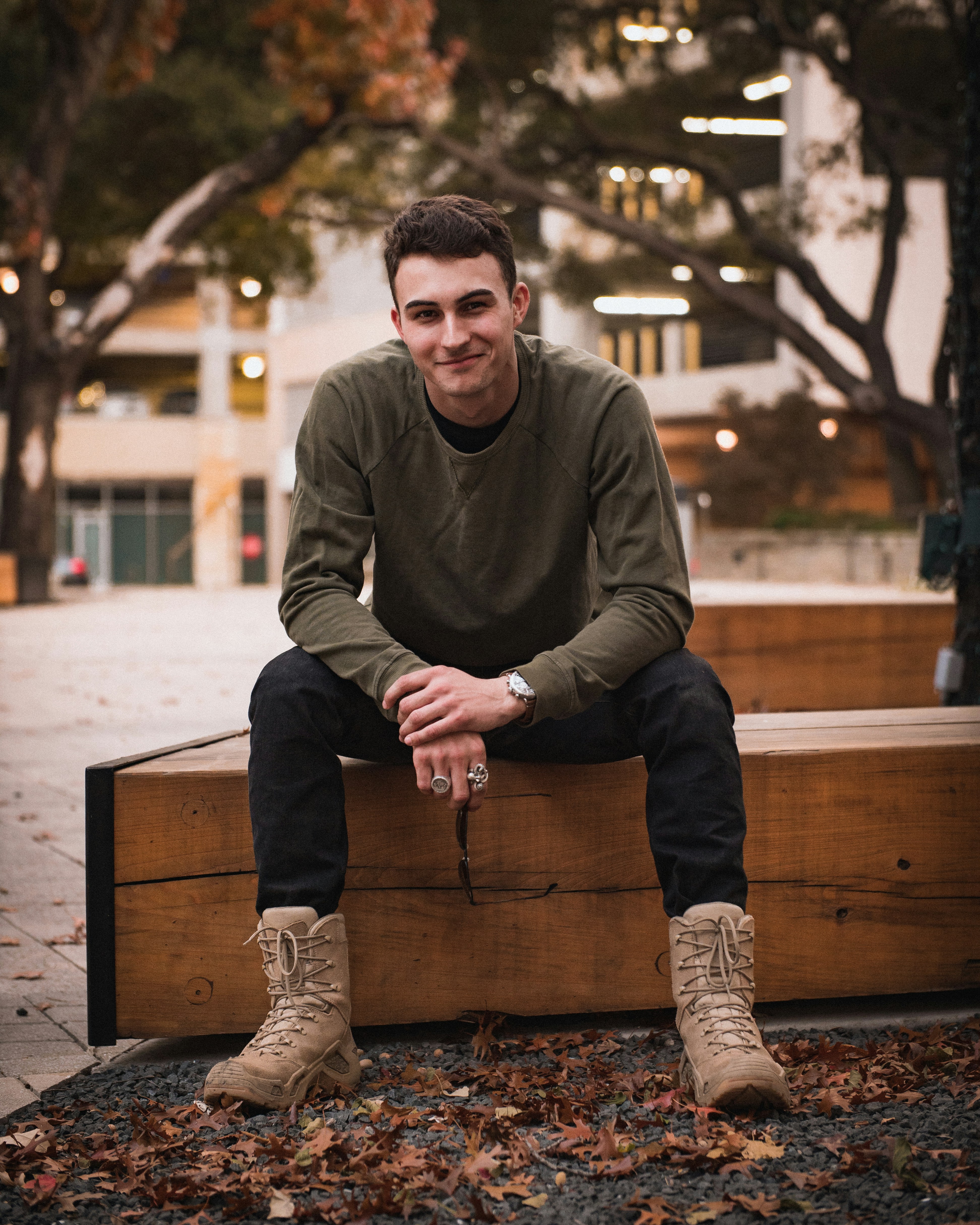 Young man seated on a wooden bench in an urban setting, exuding confidence and style. The backdrop features blurred city elements, enhancing the subject's prominence.