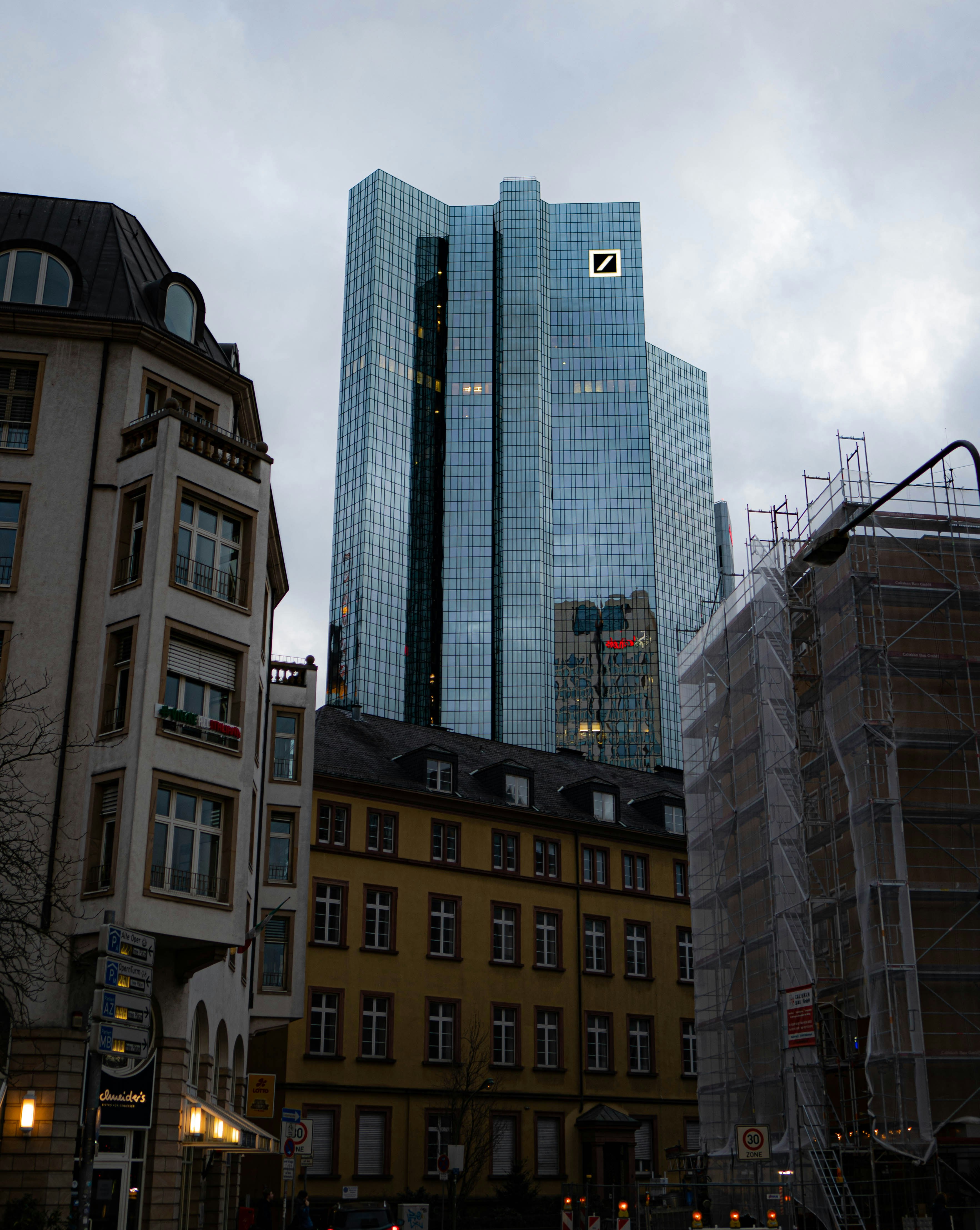 A sleek modern skyscraper reflects the surrounding historical architecture in an urban setting, with construction scaffolding hinting at ongoing development.