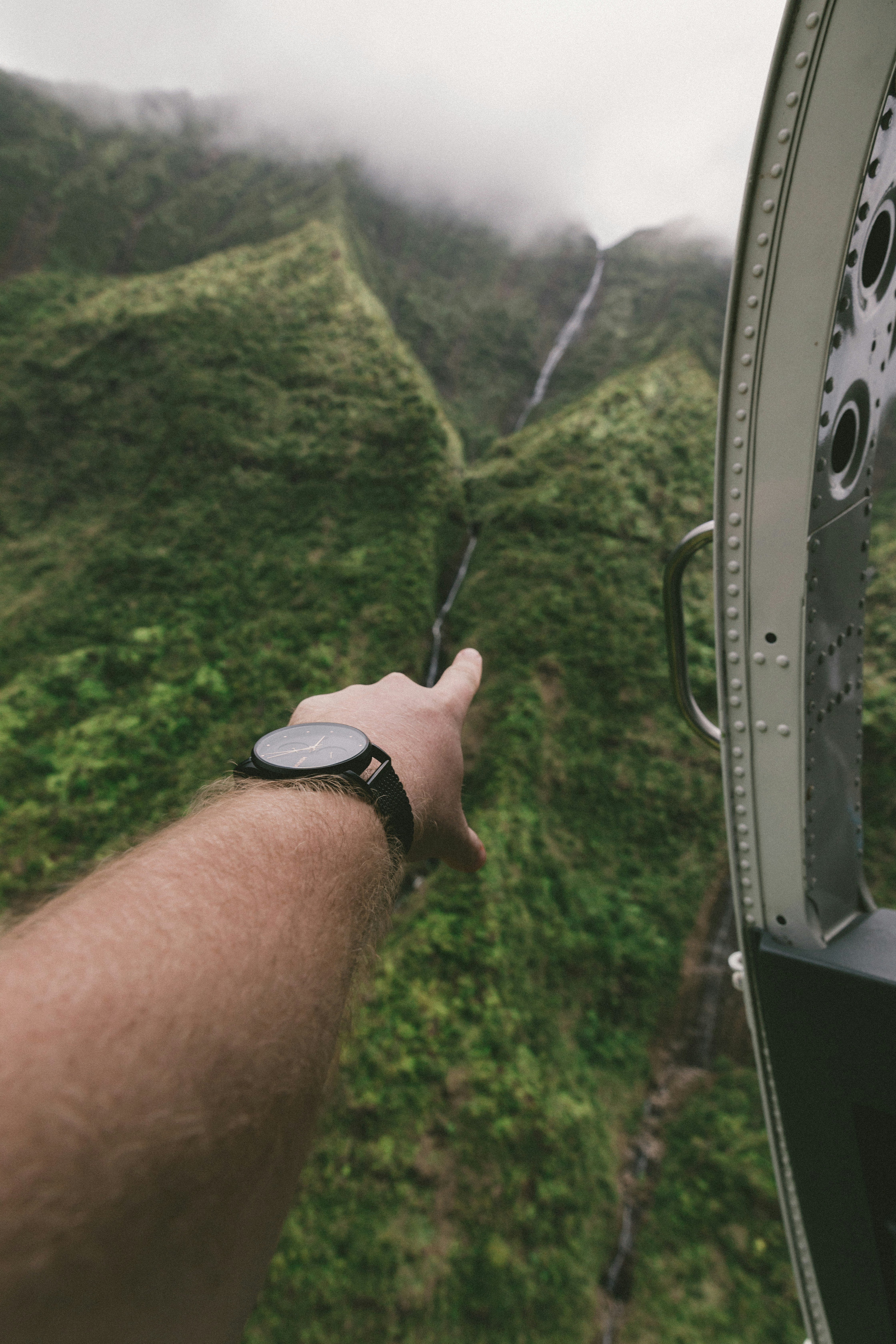 A hand with a black watch points toward a distant waterfall winding through lush green cliffs, photographed from inside a helicopter.