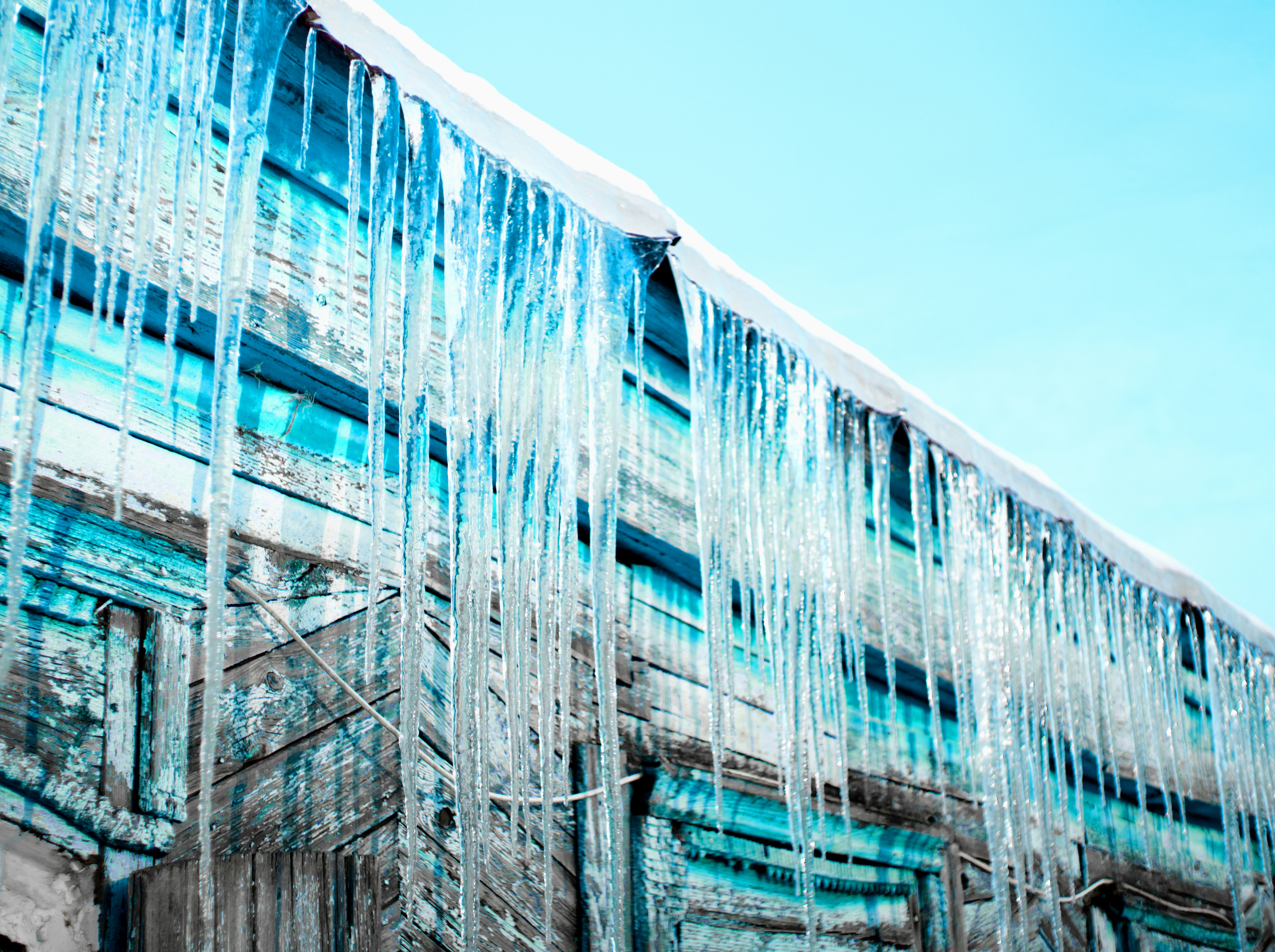 Icicles hang from a weathered building, reflecting the blue sky and showcasing the interplay of light and texture. The scene captures the beauty of winter's chill.