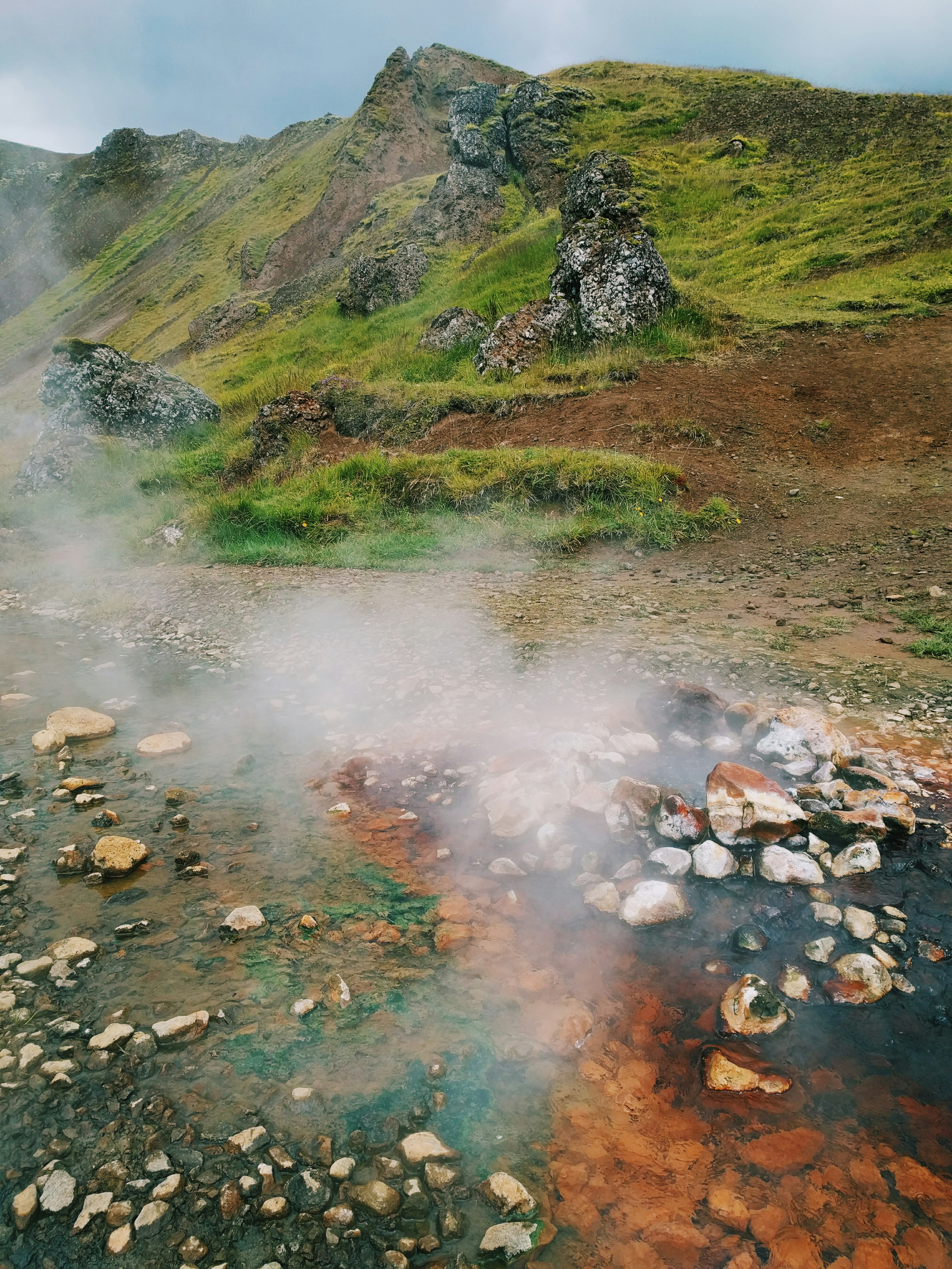 Geothermal landscape with steam rising from a rocky stream bed. Green slopes and jagged rock formations frame a solitary stone pillar.