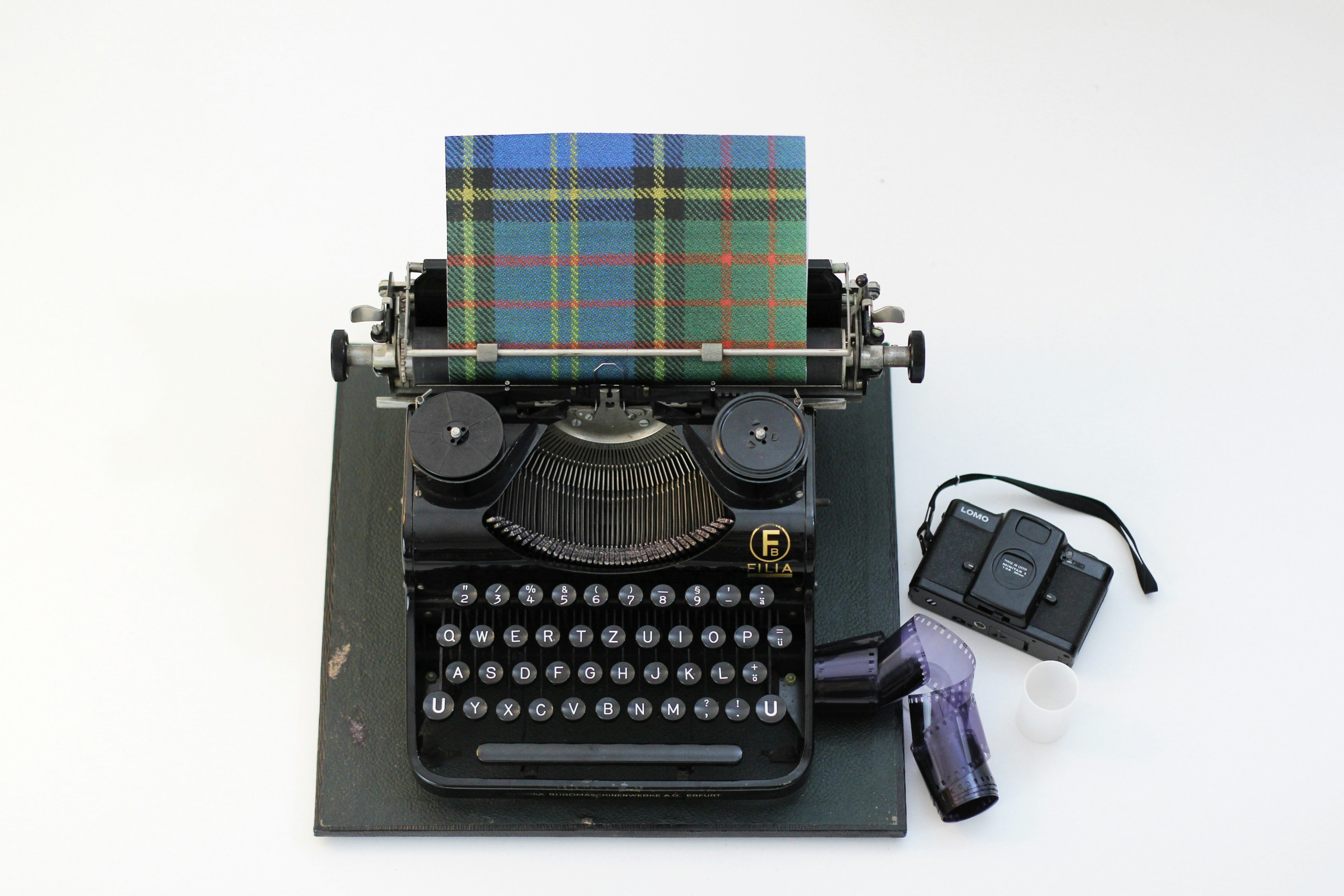 Vintage typewriter with a colorful plaid paper, accompanied by a black camera and film rolls on a white surface.