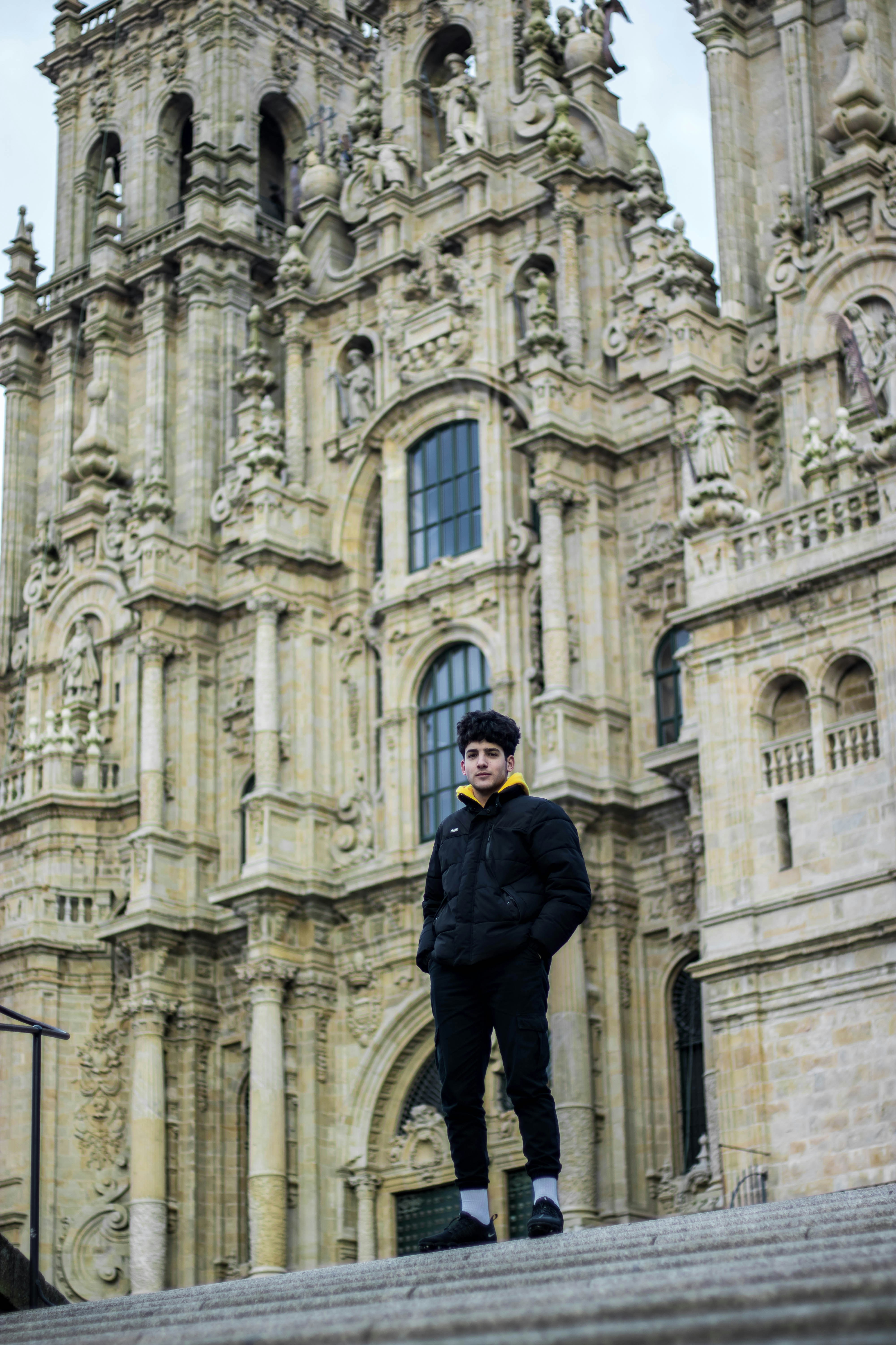 A young man stands confidently on stone steps in front of an intricately designed historical building, showcasing its detailed façade.