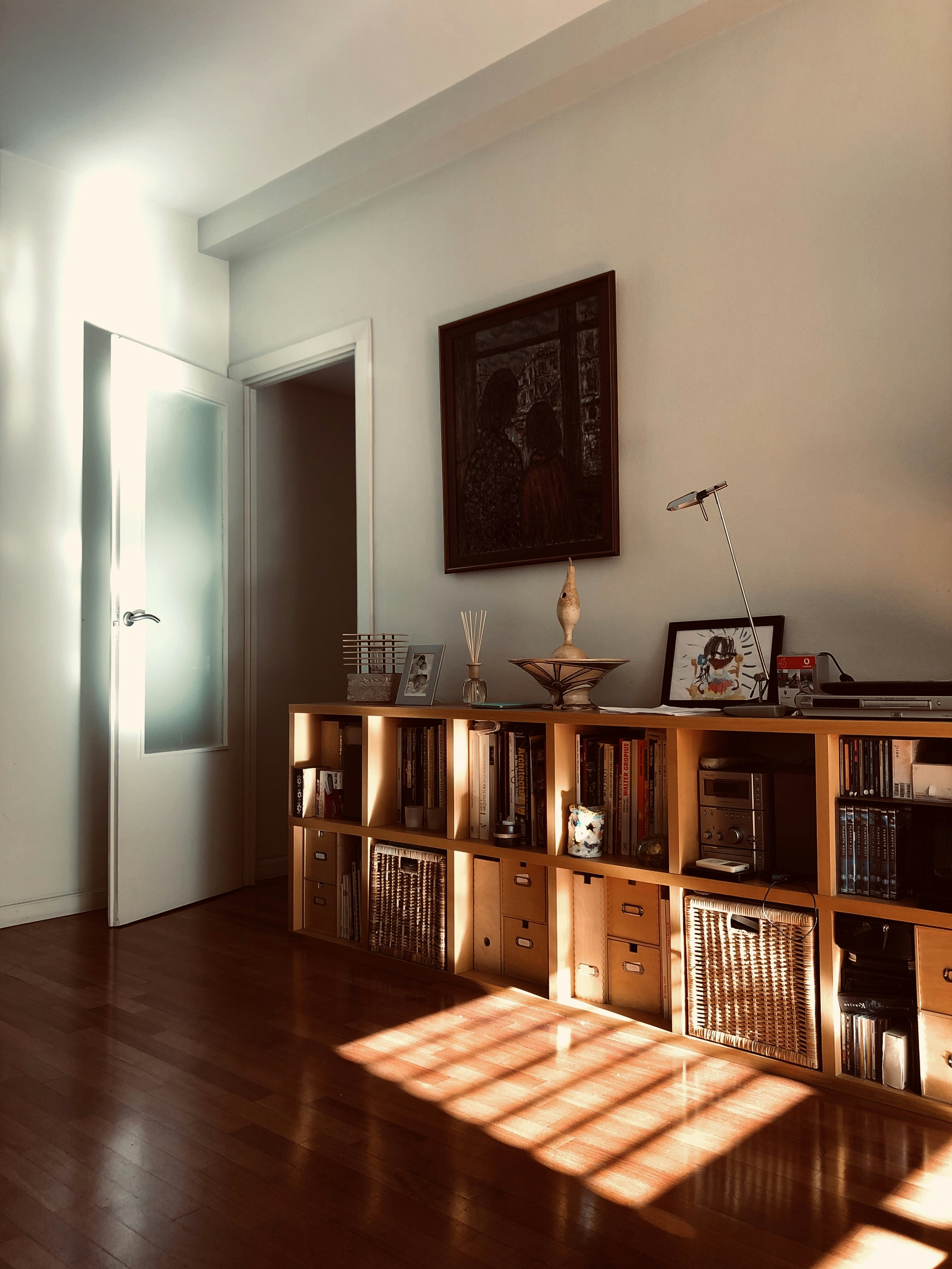 Warm sunlight filters through a doorway, illuminating a wooden bookshelf filled with books and decorative items, creating a serene indoor atmosphere.