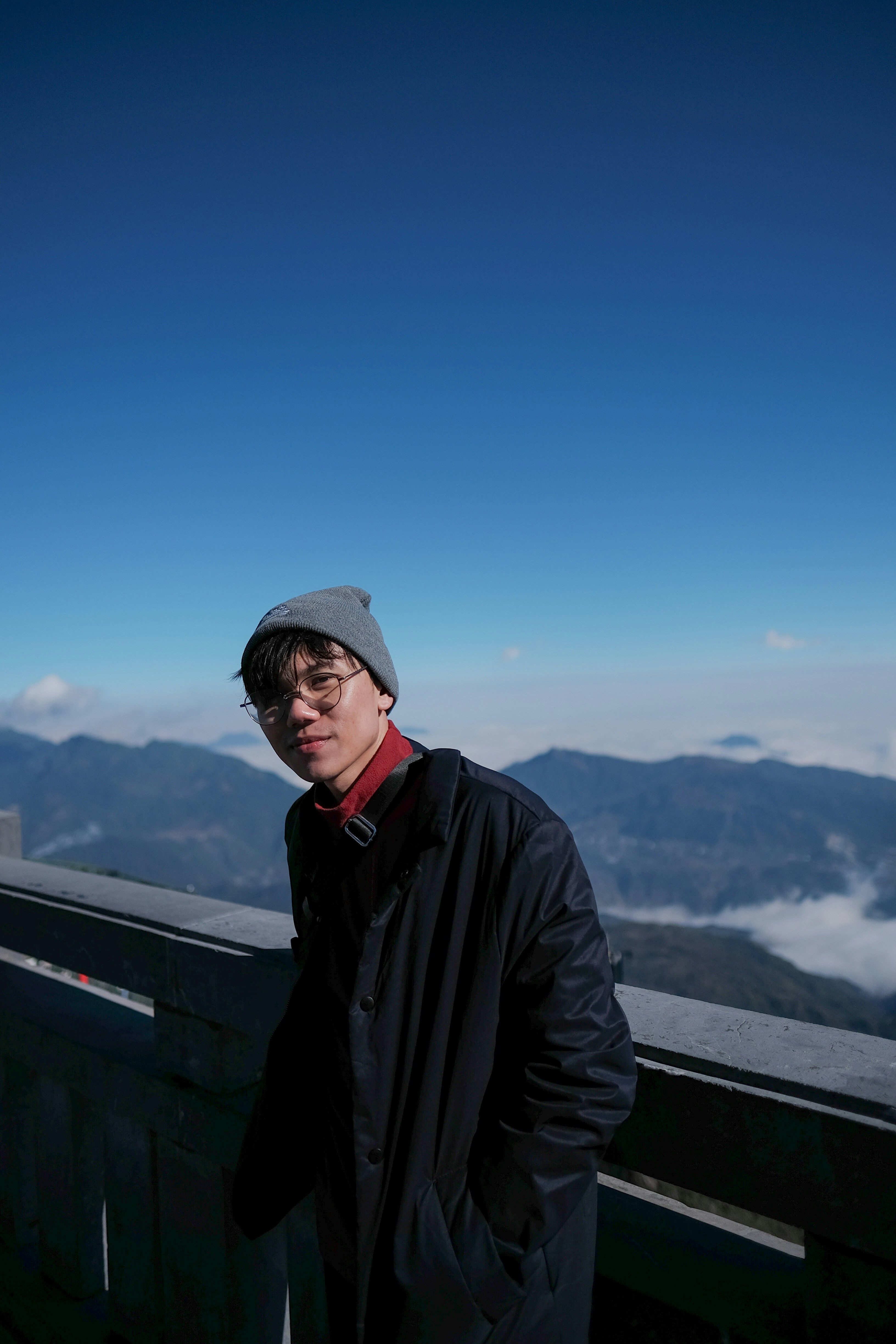 Young man in a beanie and coat stands against a backdrop of mountains and a clear blue sky, exuding a sense of adventure and tranquility.
