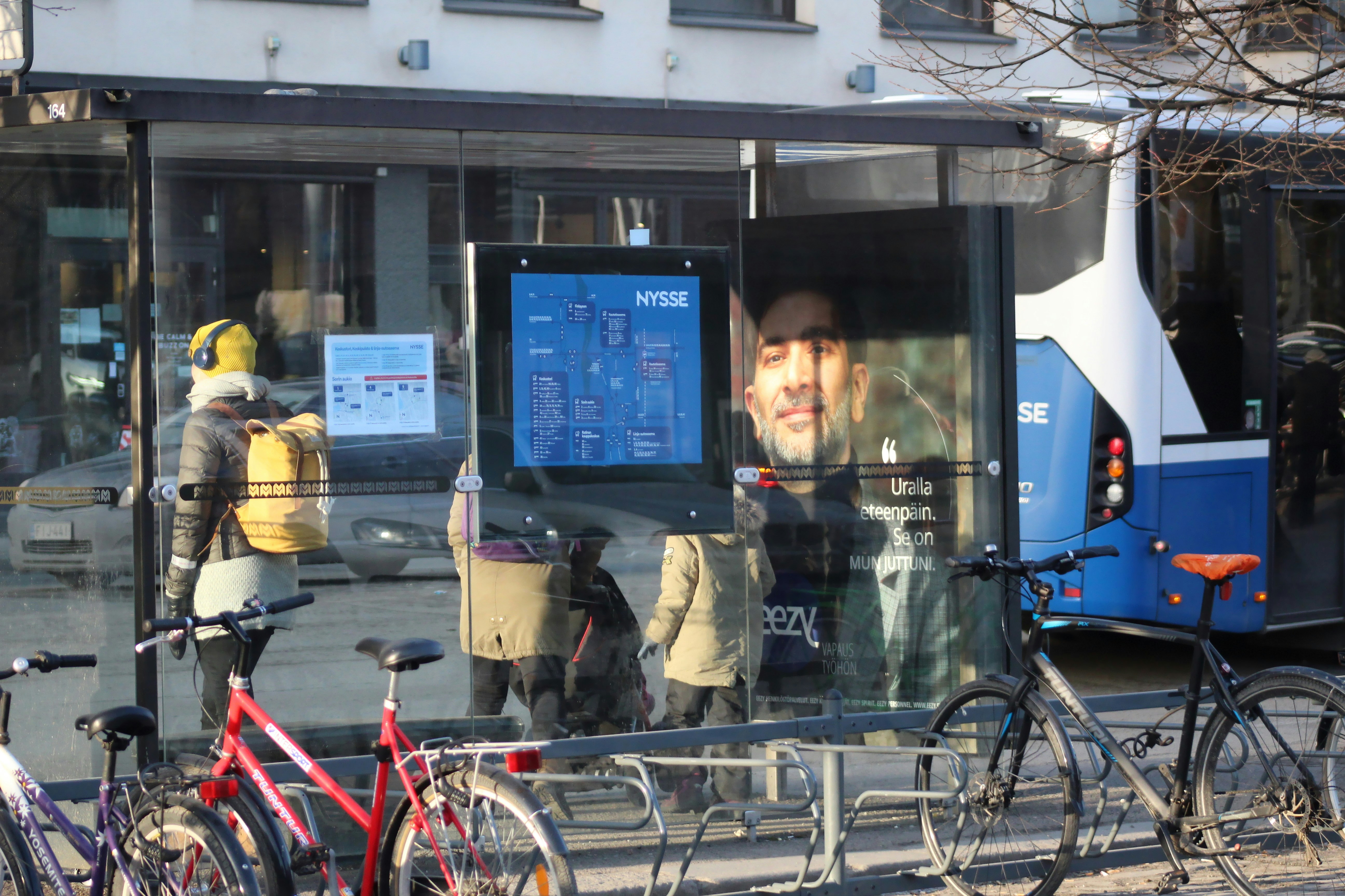 A bus stop featuring a digital display and a reflective glass showcasing a man's face, surrounded by parked bicycles in a bustling city setting.