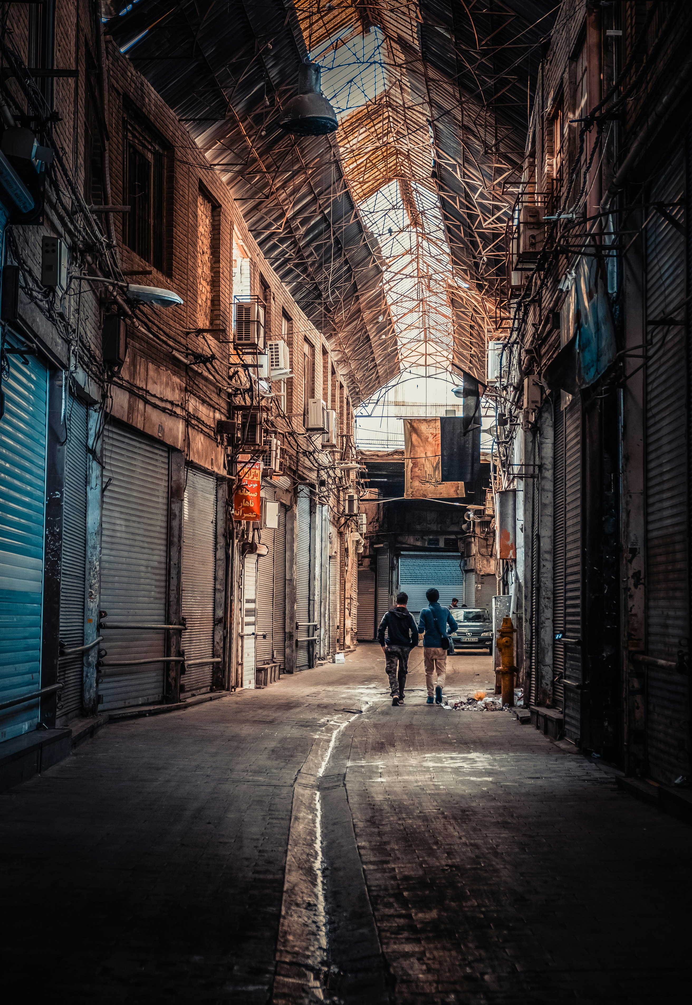 Two figures walk hand in hand down a deserted market alley, illuminated by shafts of light filtering through the roof. The scene captures a blend of nostalgia and urban decay.