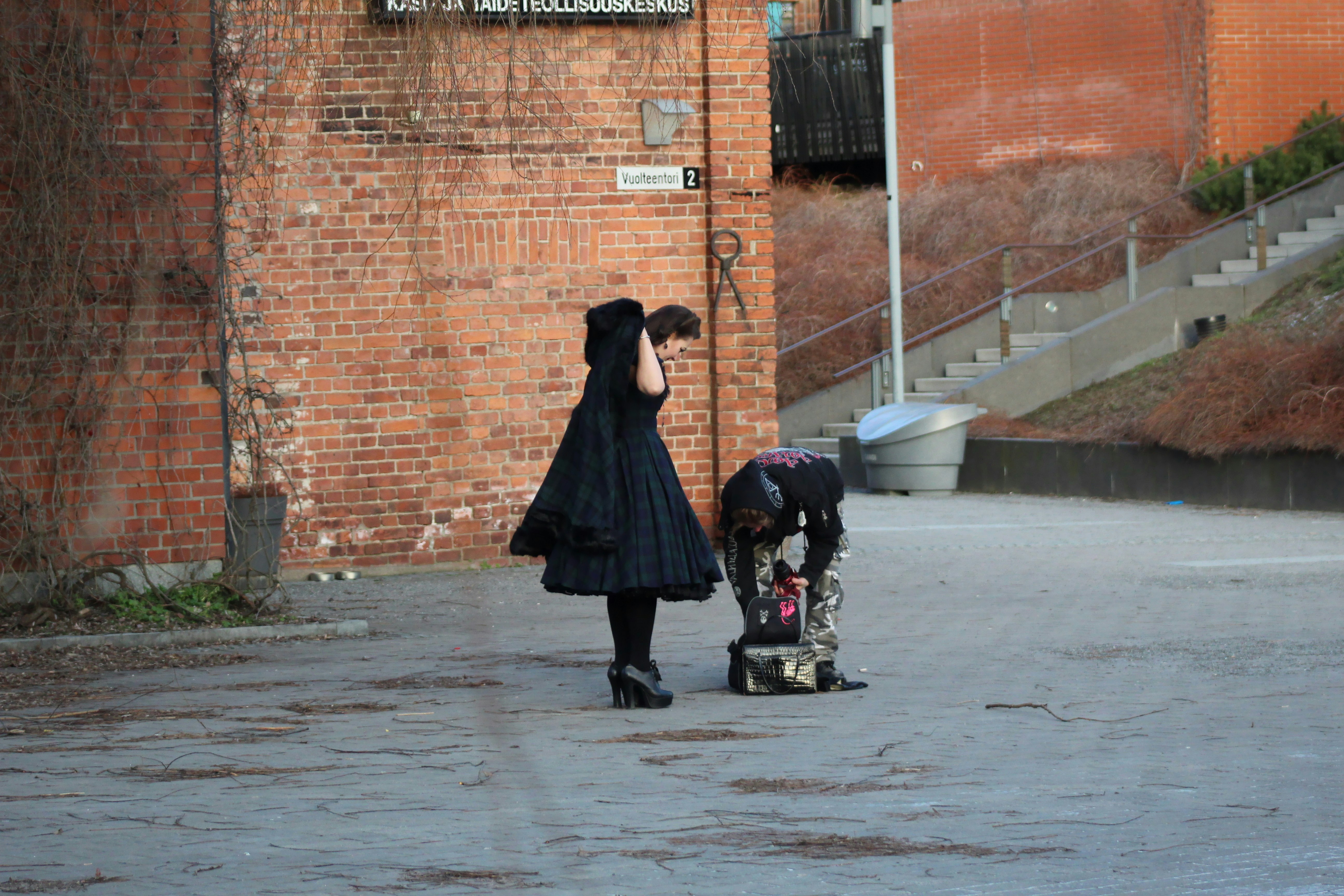 Two figures in a stylish urban setting, one adjusting their outfit while the other prepares equipment. A blend of fashion and street life.