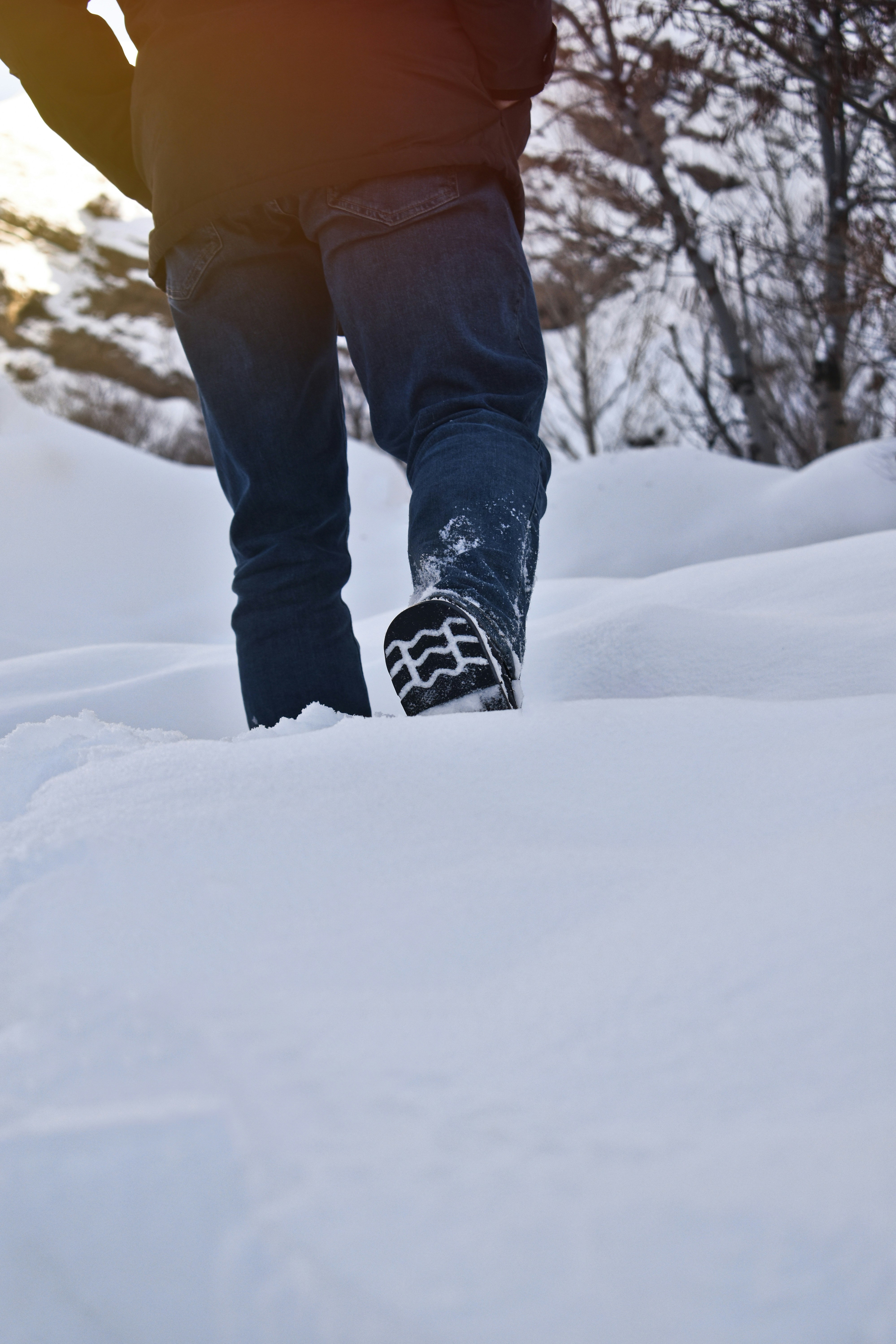 Person in dark clothing walking through deep snow with sunlight filtering through trees.