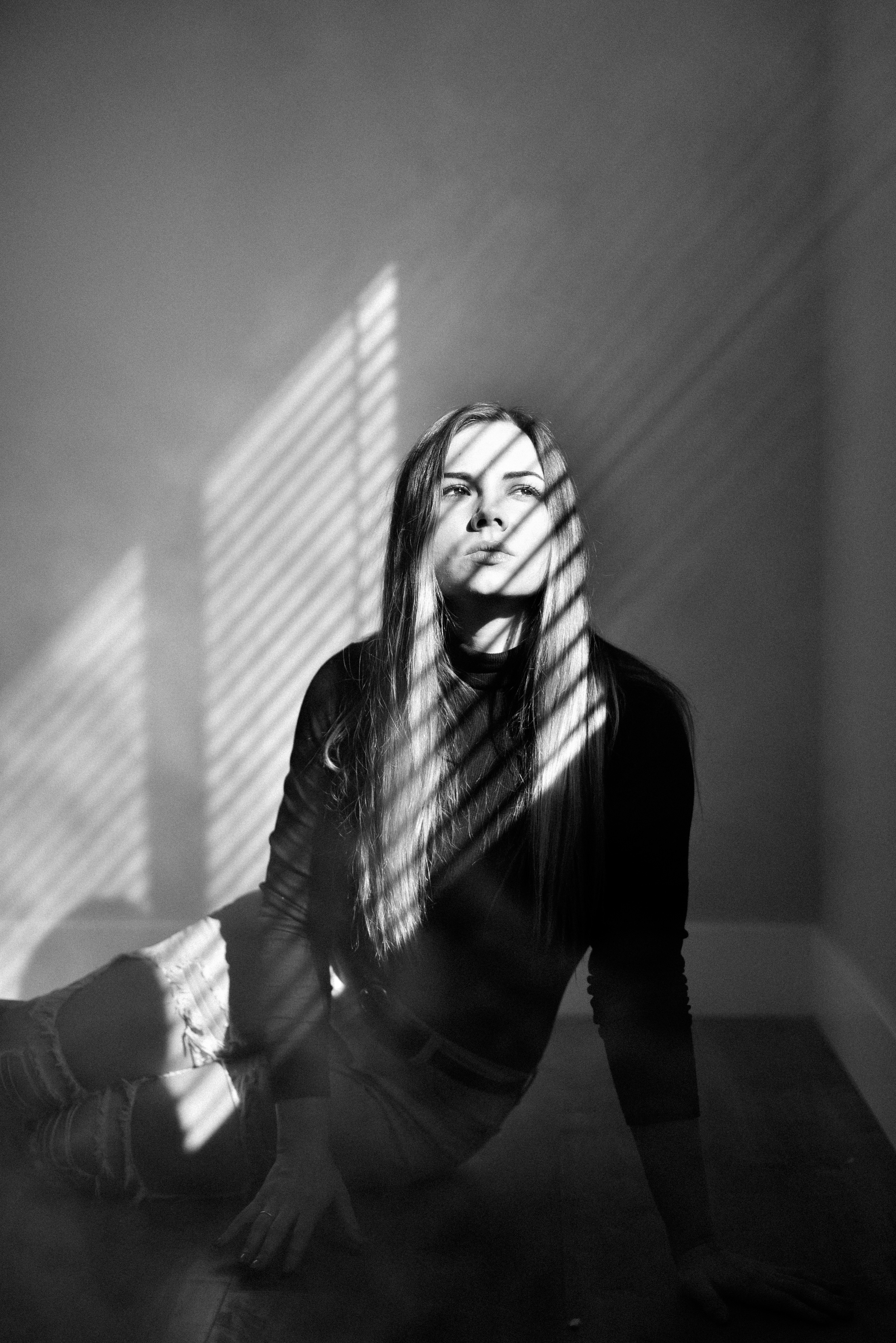 A woman sitting on the floor, illuminated by slanted light casting dramatic shadows across her face and body. The monochrome tones enhance the mood of introspection.
