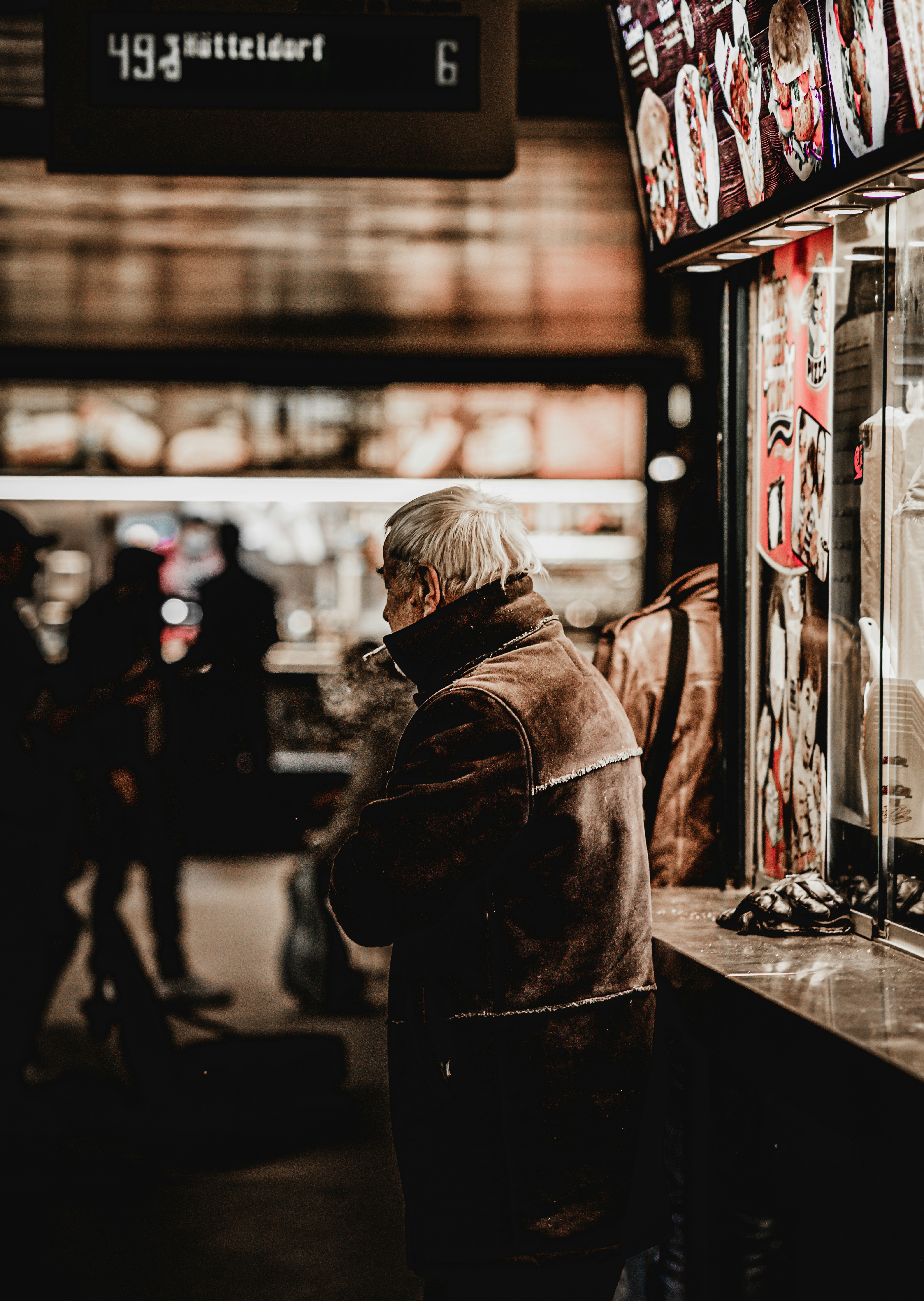 An elderly man stands thoughtfully in a bustling market, surrounded by vibrant food displays and the energy of passersby.