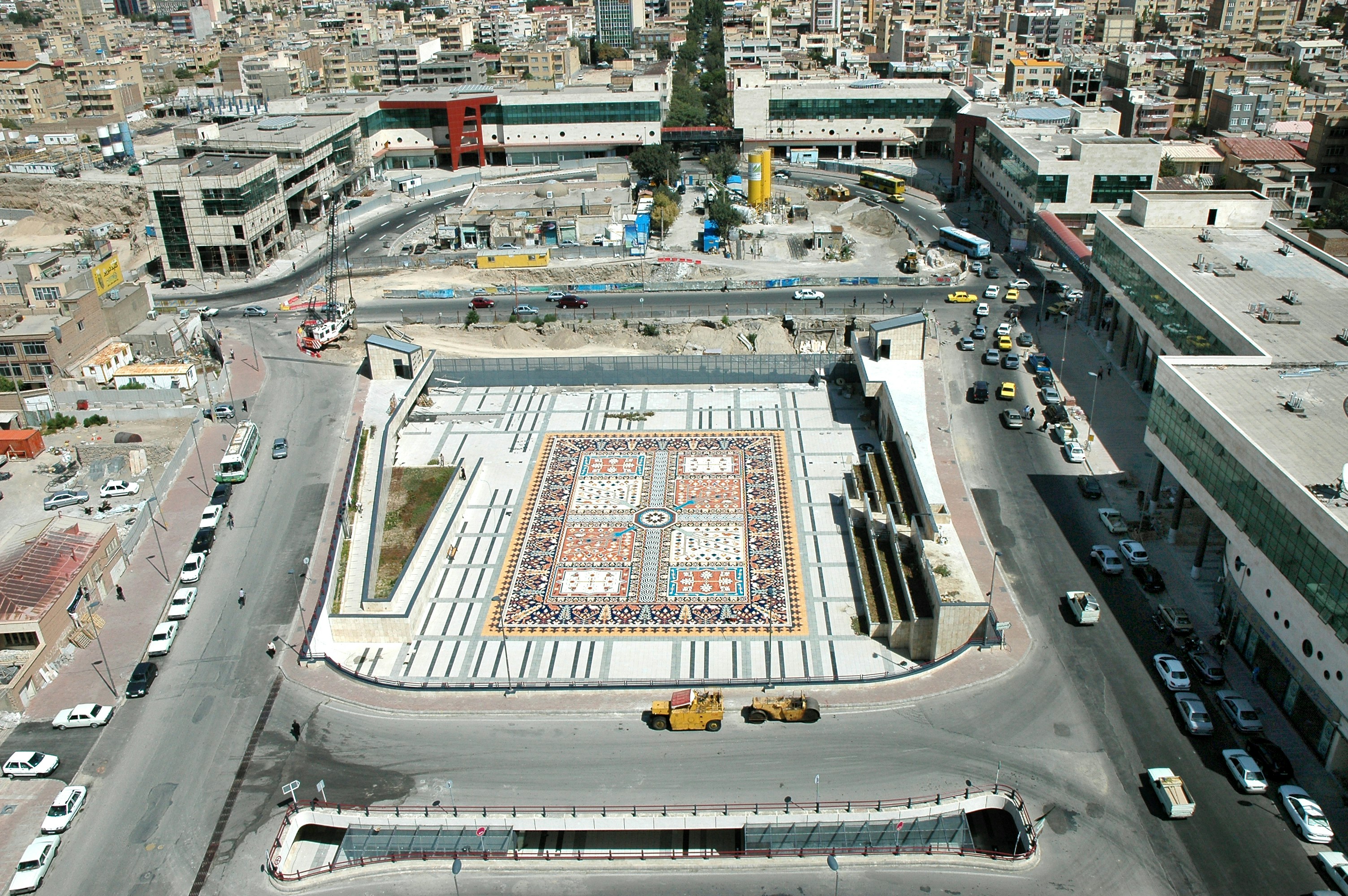 Aerial view of a large, symmetrical city square surrounded by buildings and roads, with construction equipment visible.
