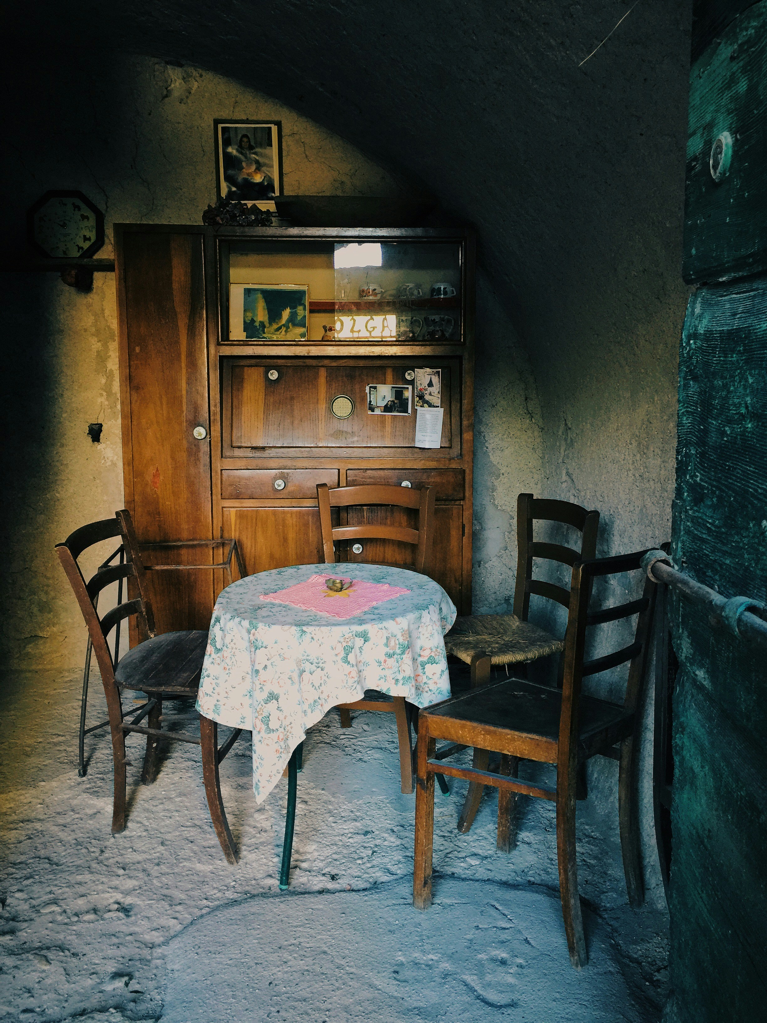 Cozy interior featuring a round table with a floral tablecloth surrounded by wooden chairs, illuminated by soft light filtering through the room. A vintage cabinet holds cherished memories on the wall.