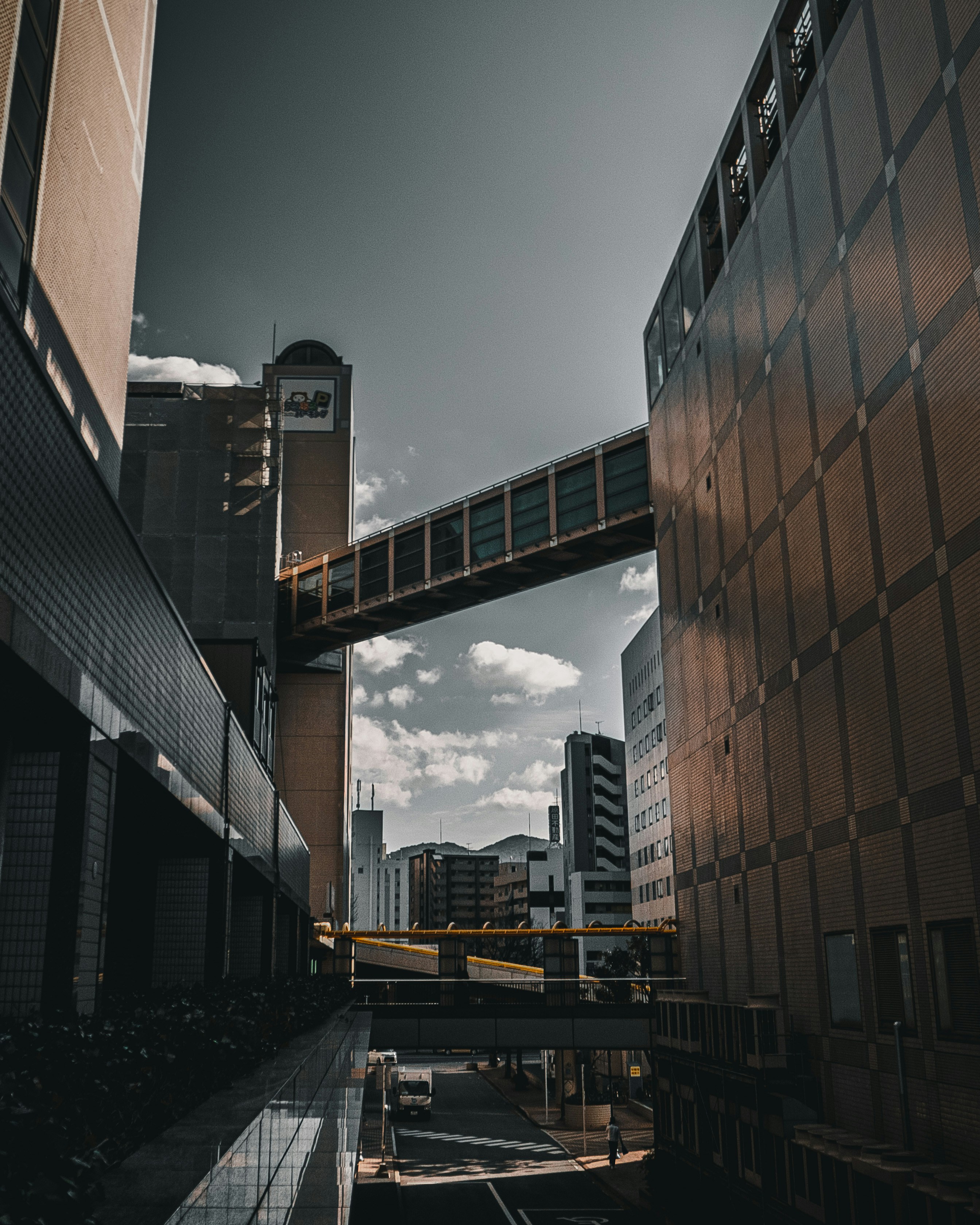 A modern pedestrian bridge connects two buildings, framed by urban architecture and scattered clouds above. The scene captures the blend of functionality and design in a bustling city.