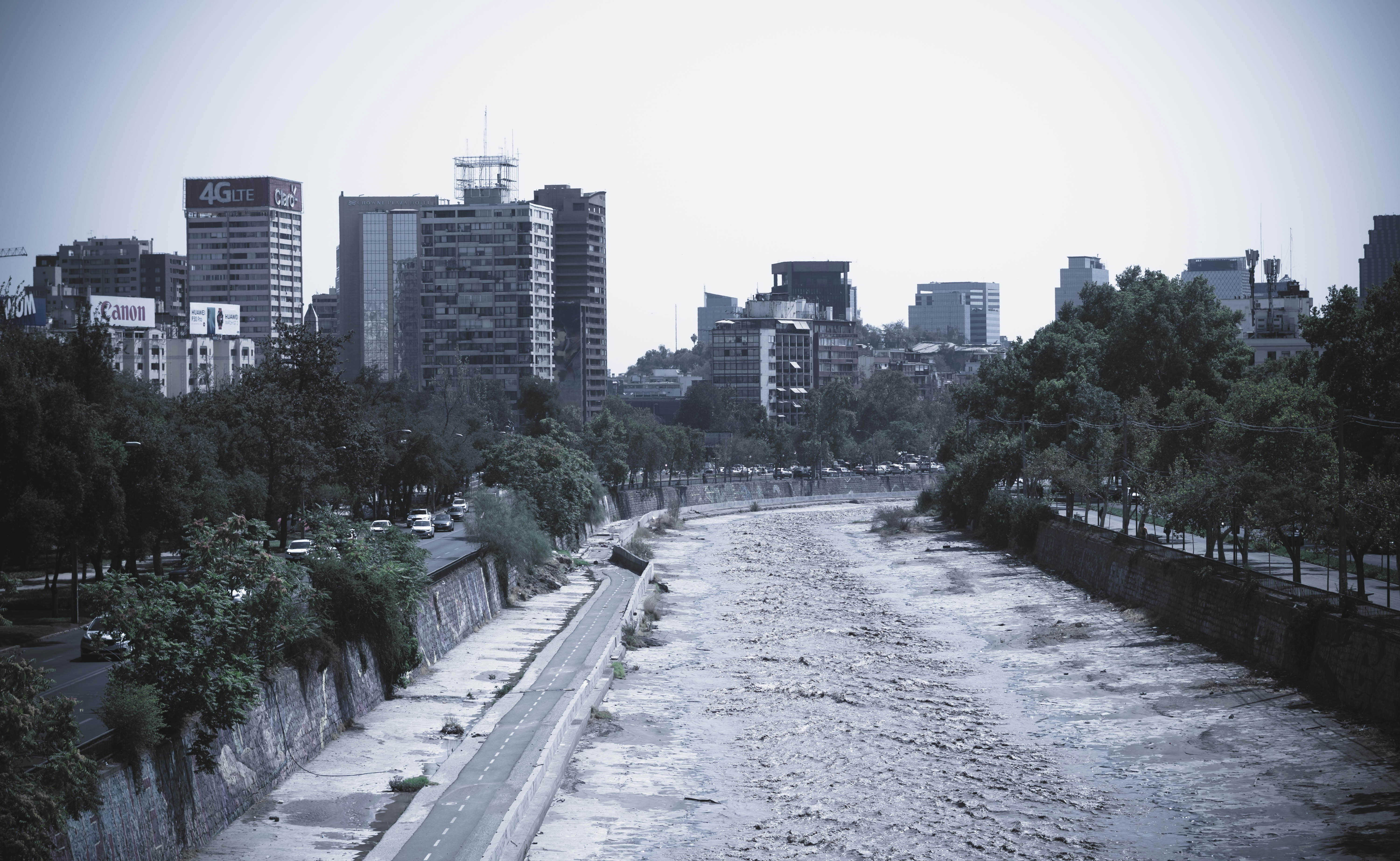 Gray concrete riverbed flanked by green trees and tall buildings under a bright sky.
