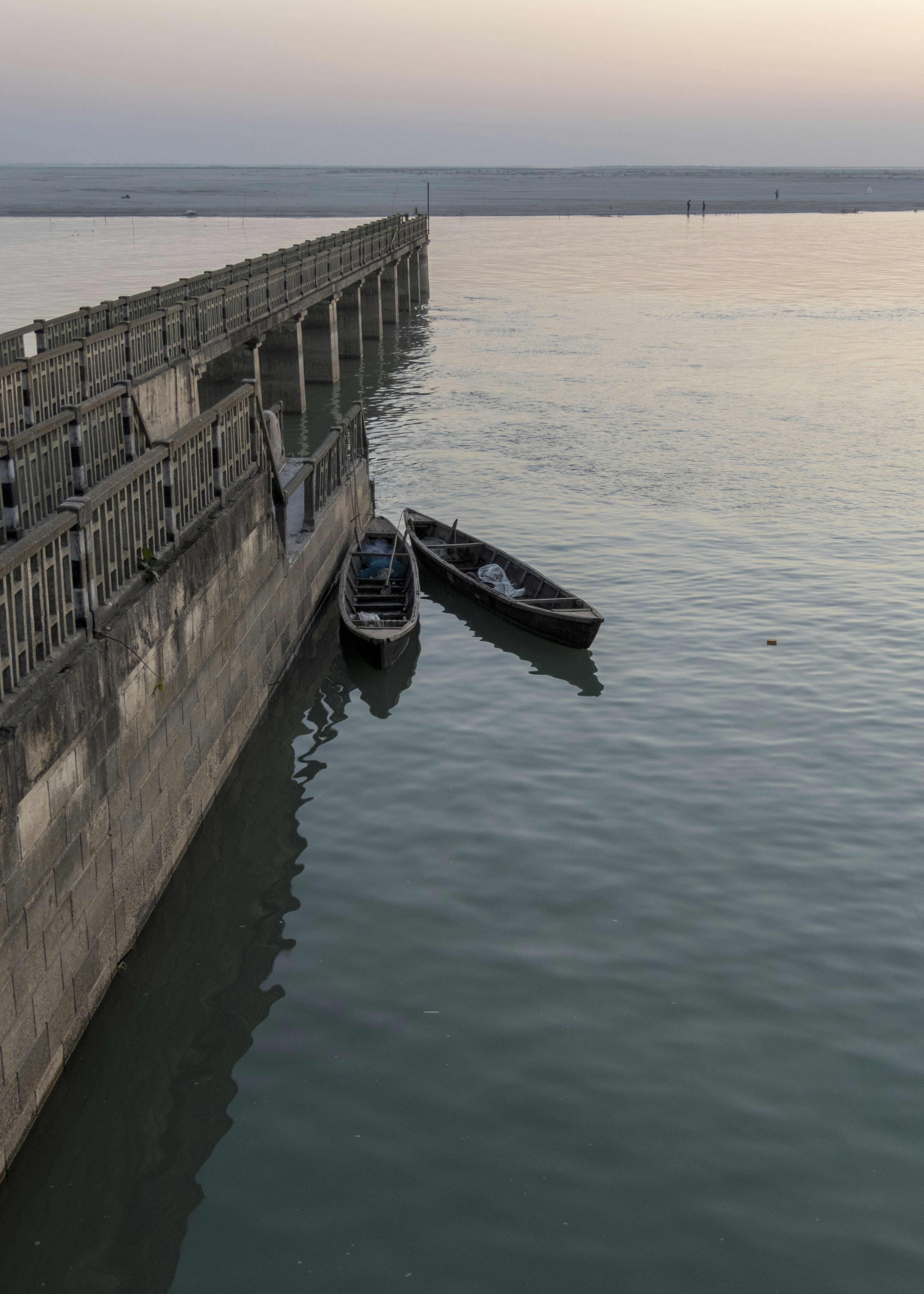 Two small boats moored along a weathered concrete pier, with calm water and a distant horizon at dusk.