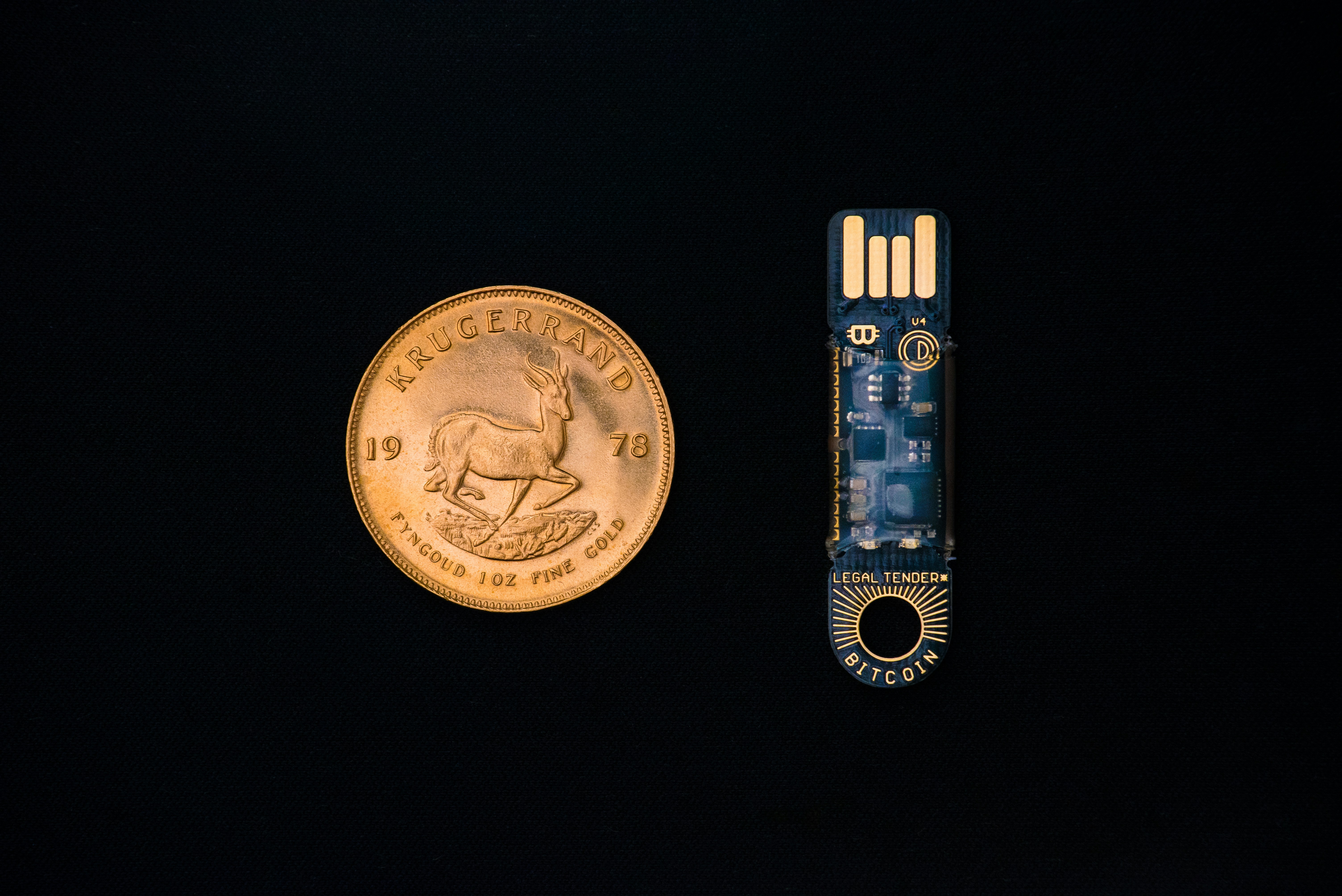 A gold Krugerrand coin positioned next to a sleek Bitcoin hardware wallet against a black background.