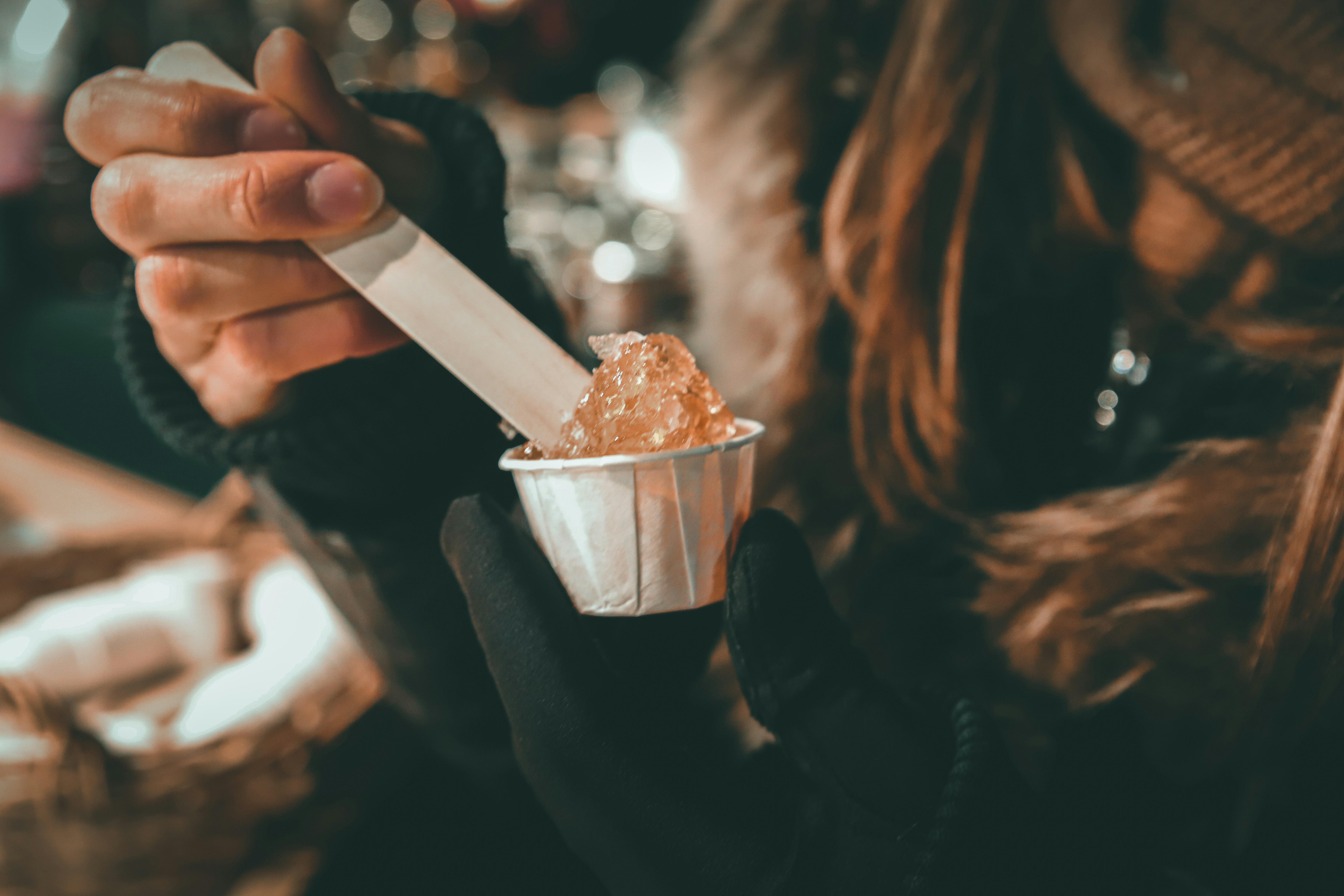 A hand holding a spoon poised over a small cup of golden syrup, surrounded by a warm, festive atmosphere.
