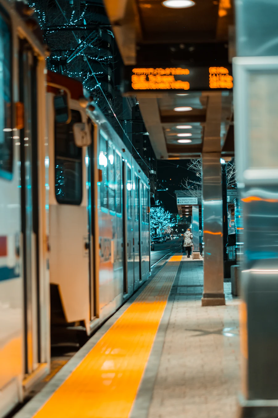 A transit platform at night with a train along the left side, glowing orange signage, and a lone commuter in the distance. Cool blue reflections contrast with the warm lights overhead.