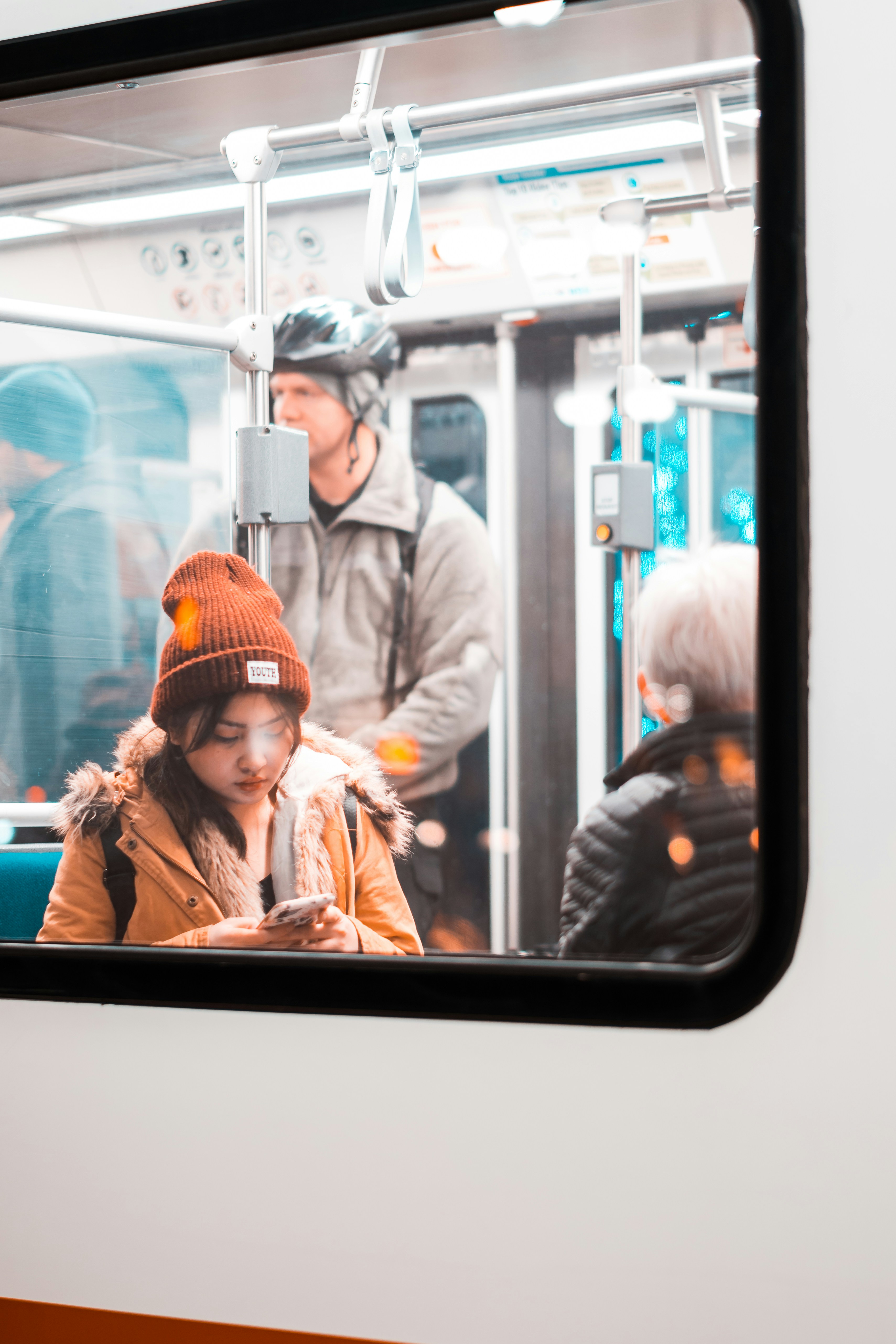 A young woman focused on her phone sits inside a train compartment, framed by the window, as other passengers appear in the background. The scene captures a slice of urban life during a commute.