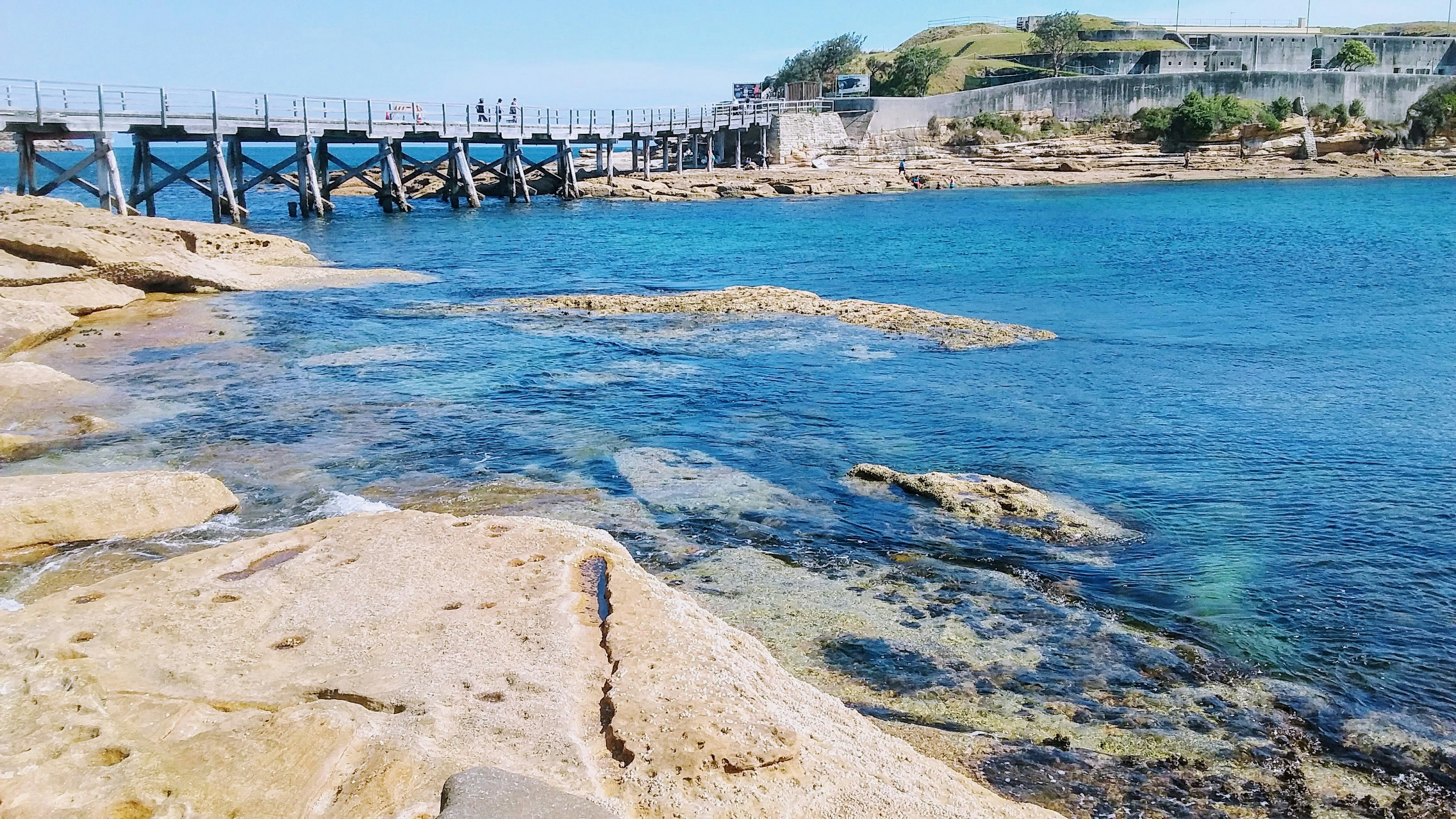 Rocky shoreline meets tranquil turquoise waters, with a wooden pier extending into the distance. A lush hillside frames the scene.