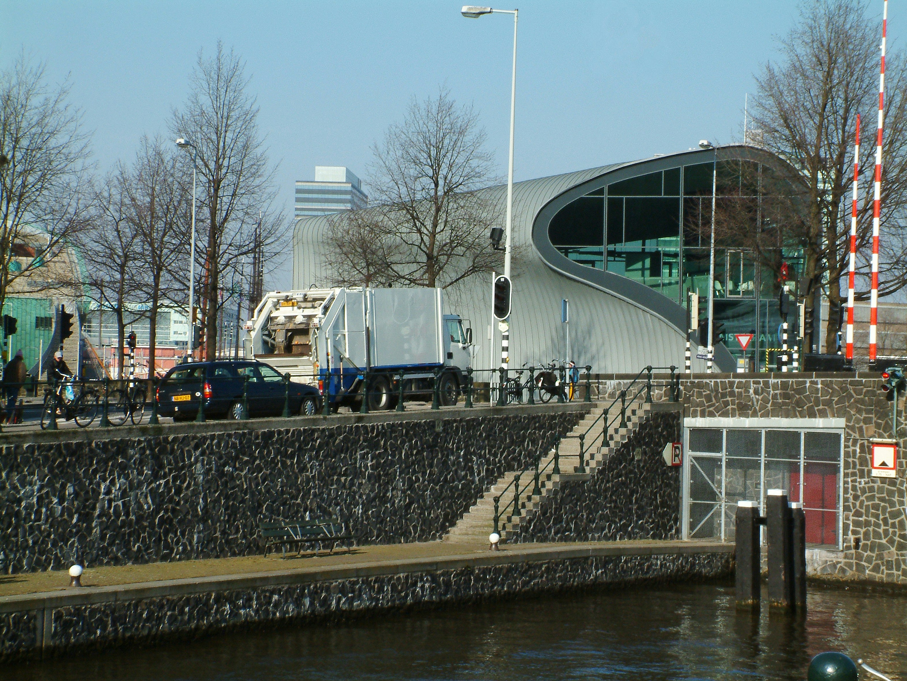 Modern building with a curved facade alongside a canal, framed by bare trees and urban infrastructure.
