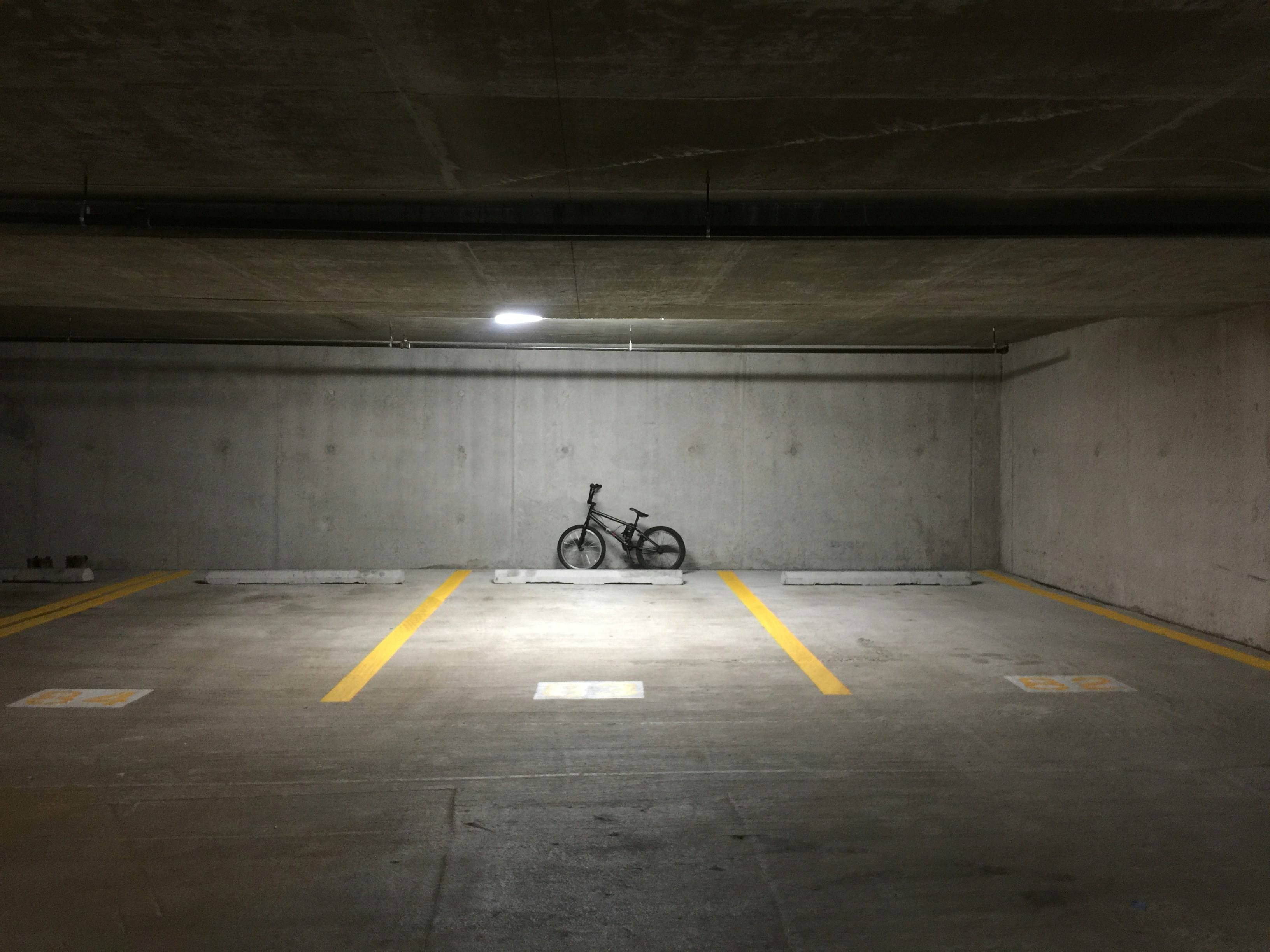 A lone bicycle rests against a concrete wall in an empty parking garage, illuminated by overhead lighting. The stark lines of yellow parking spaces contrast with the gray surroundings.